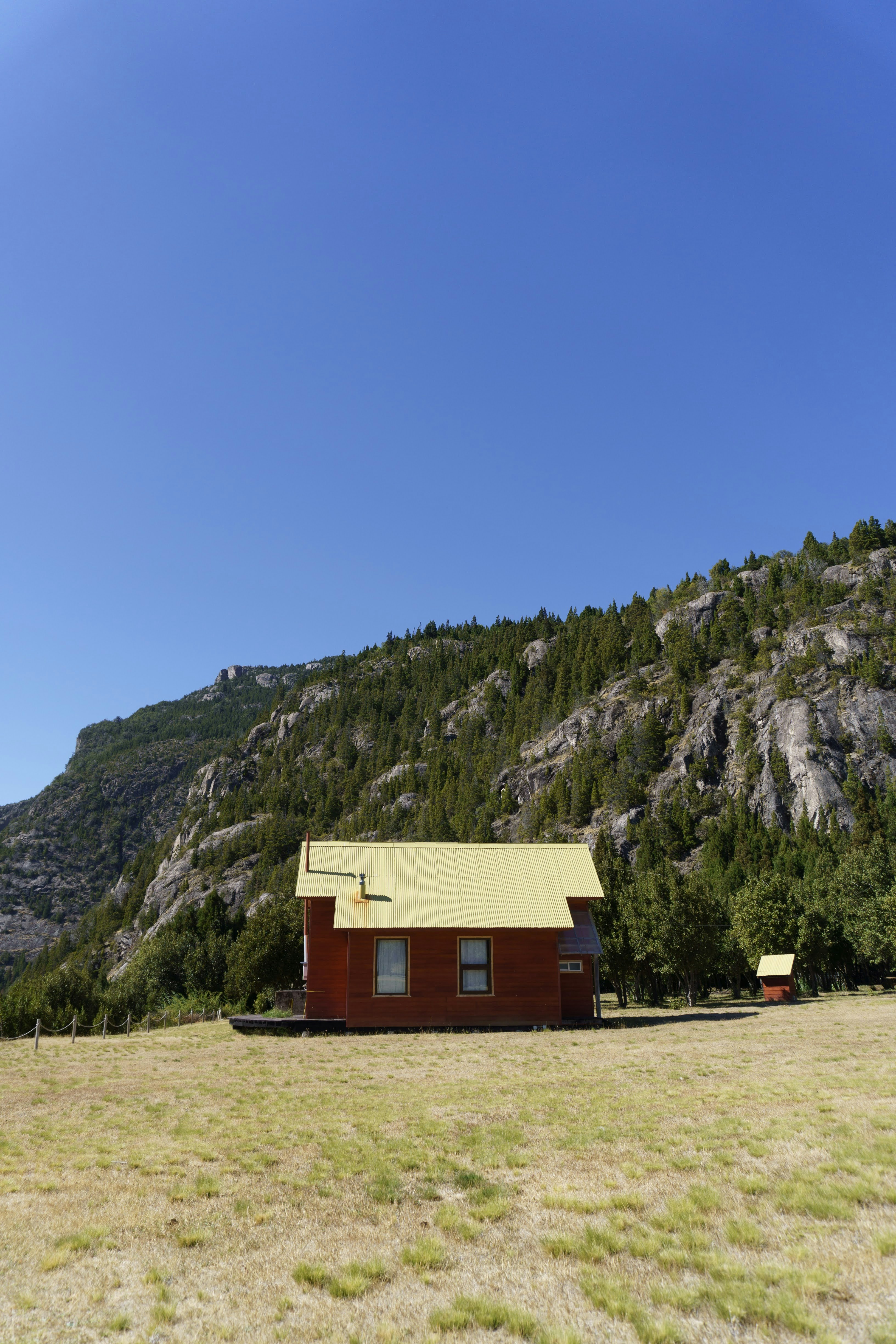 A quaint wooden cabin with a yellow roof nestled in a vast meadow, surrounded by towering mountains and lush greenery.