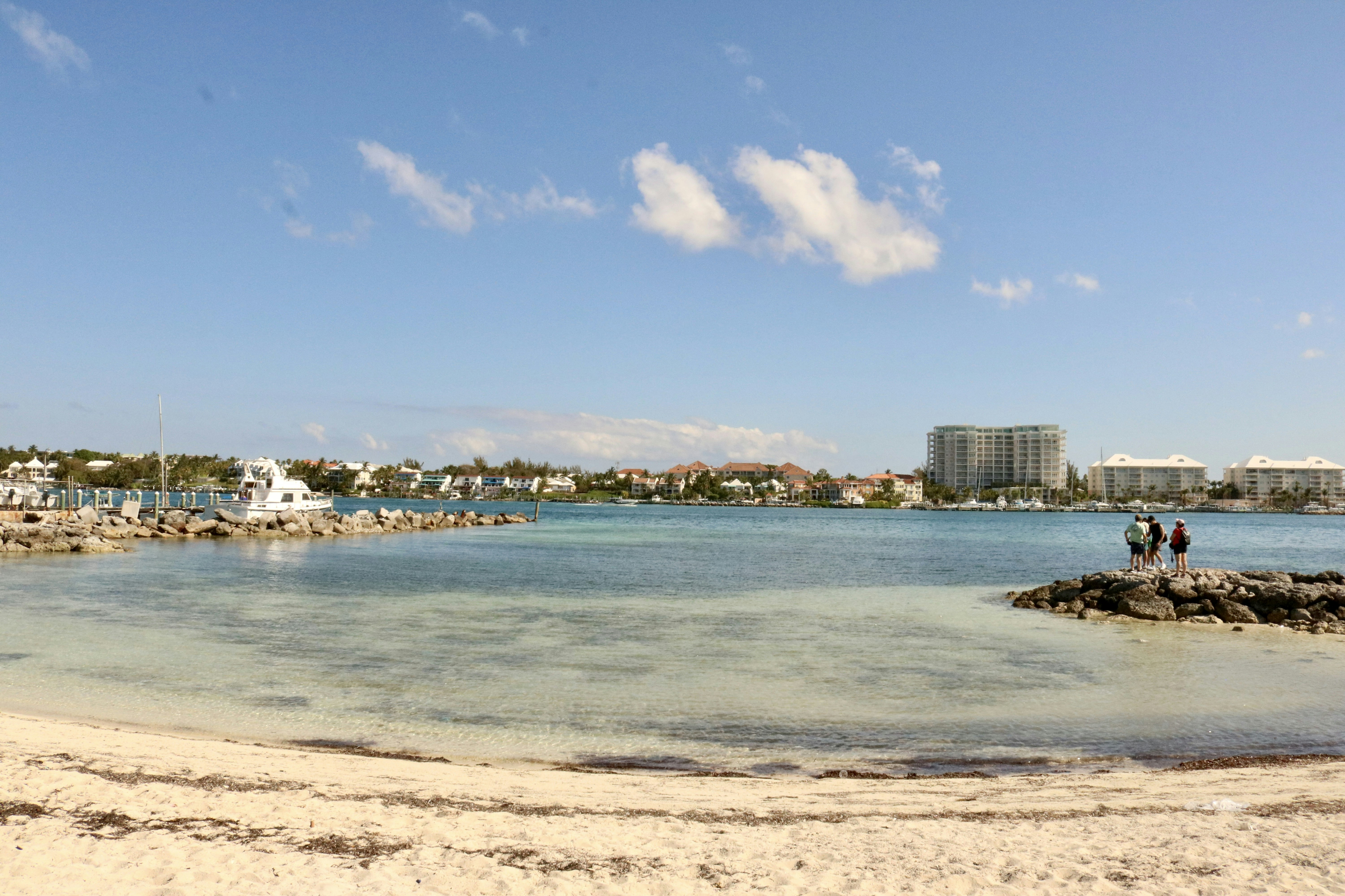 Tranquil beach scene featuring a calm bay with boats and people enjoying the waterfront. The clear blue sky complements the serene atmosphere.