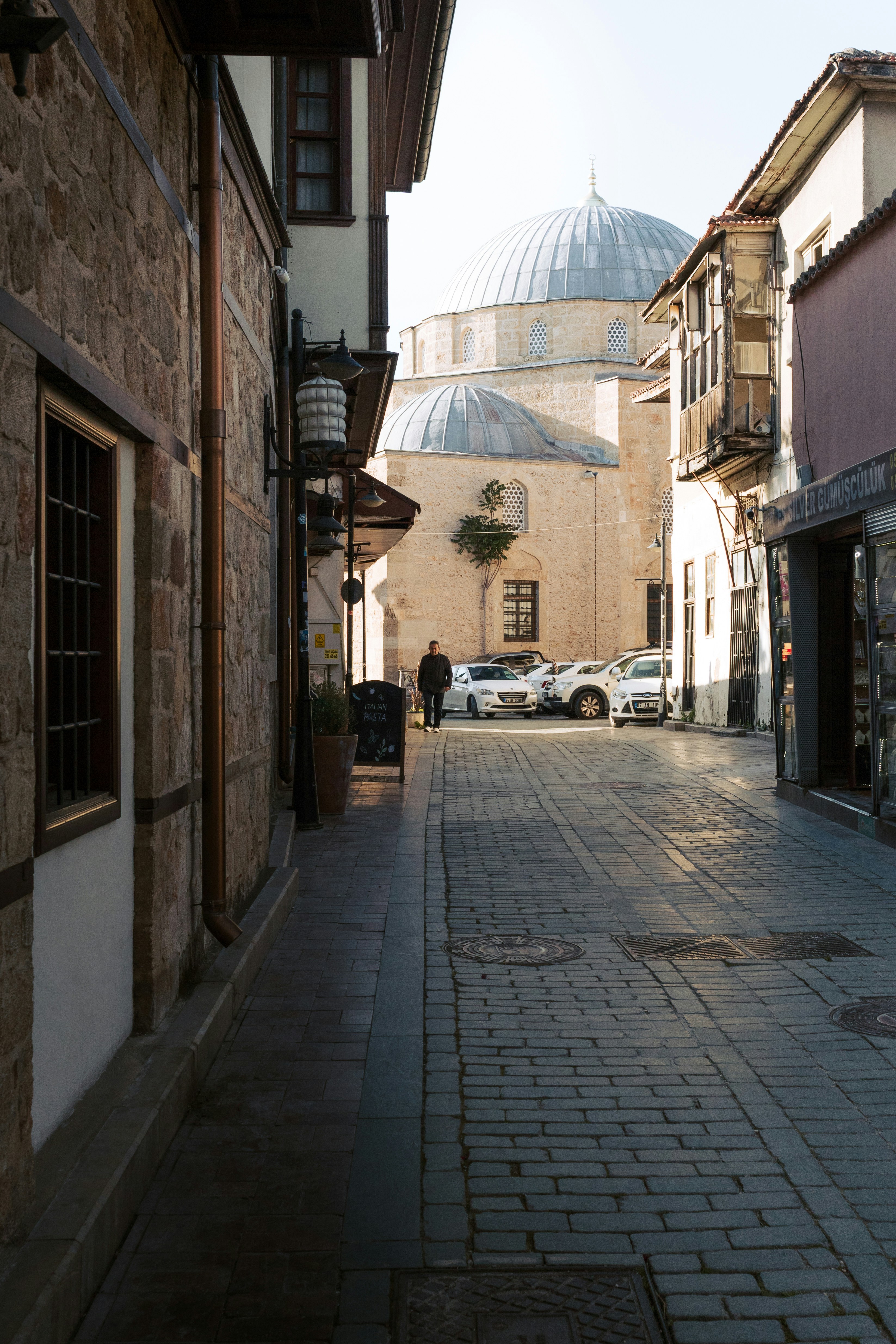 A narrow cobblestone street flanked by historic stone buildings, leading to a domed structure in the background. A solitary figure walks, adding a sense of scale and life to the scene.