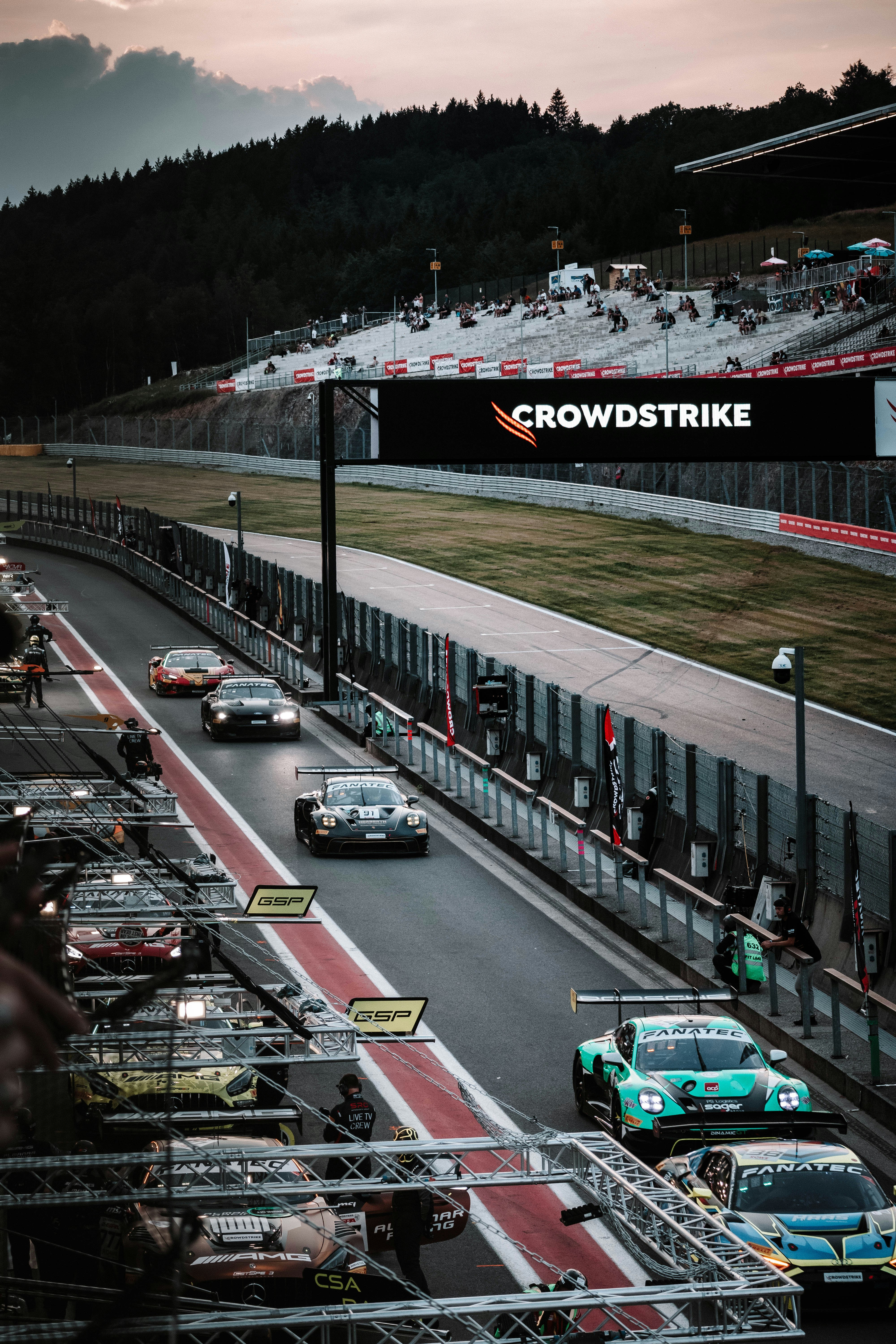 Race track action with multiple cars approaching the finish under a large CrowdStrike banner; pit walls, grandstands, and a distant forest backdrop create depth and context.