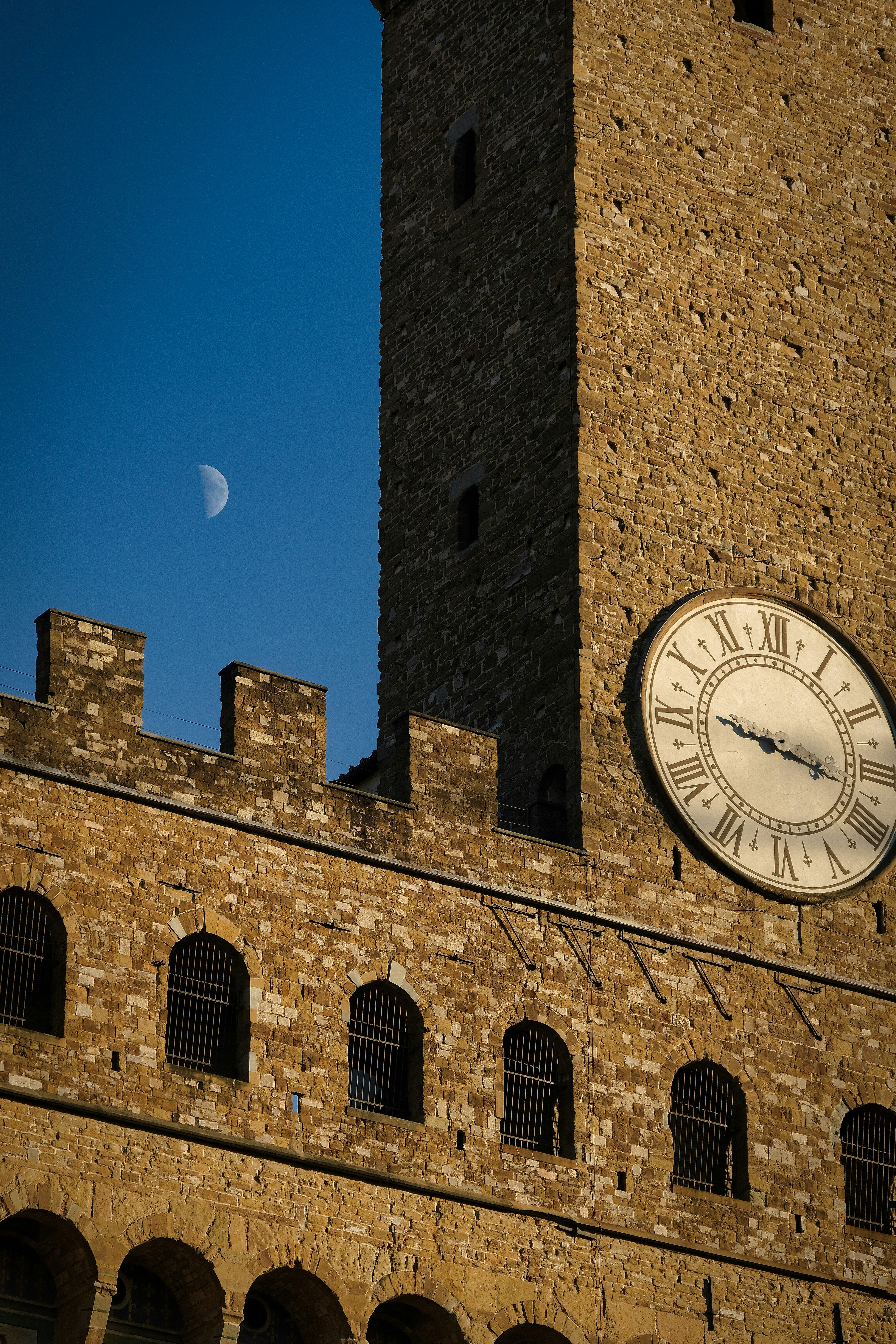 A late afternoon moon hovers over the medieval tower of Palazzo Vecchio in Florence, Italy, as golden sunlight grazes the textured stone facade and the massive Roman numeral clock marks the passing hour. The juxtaposition of celestial rhythm and man-made timekeeping invites reflection on permanence and transience.