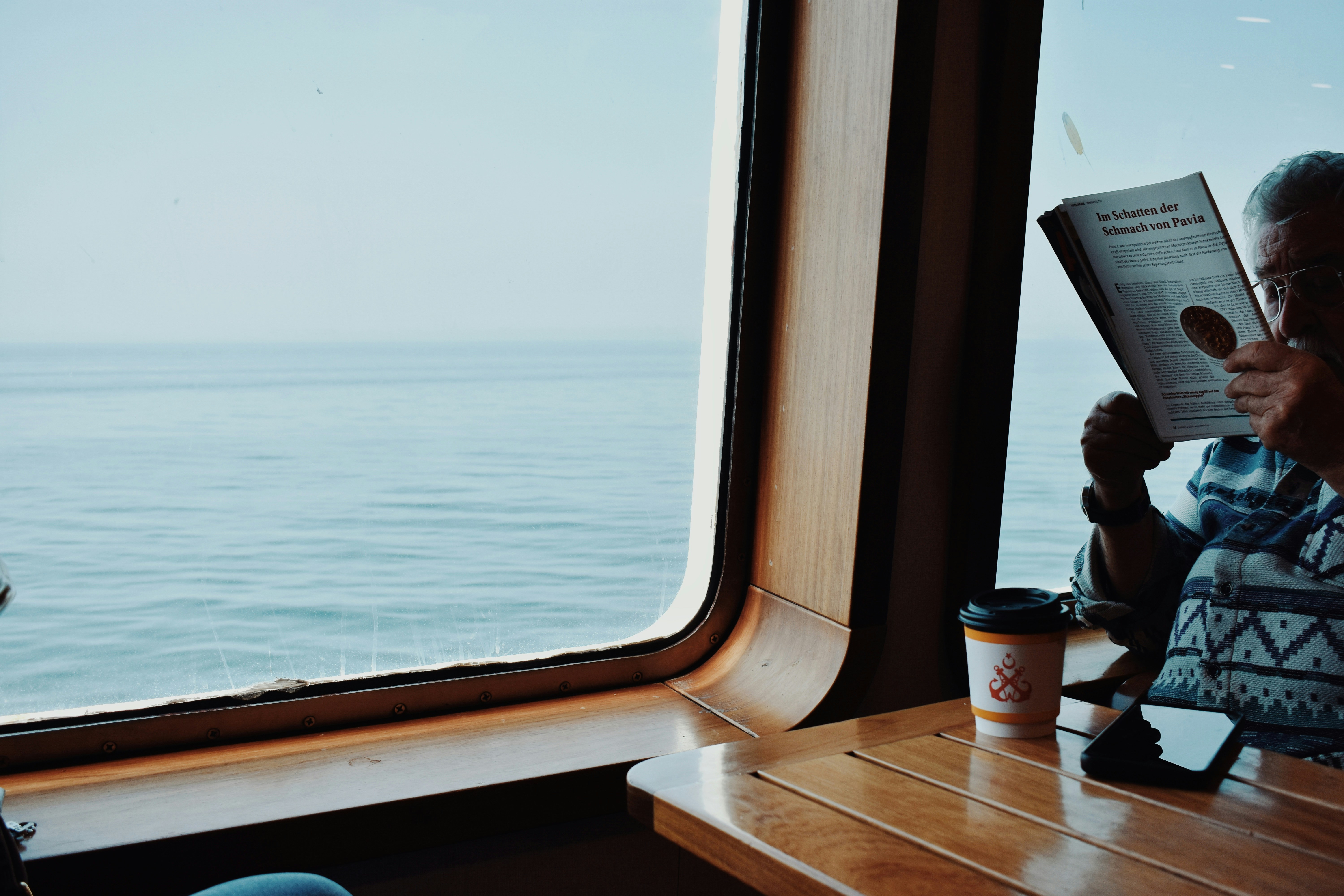 Person reading a book by a large window overlooking a calm sea, with a coffee cup on the table.