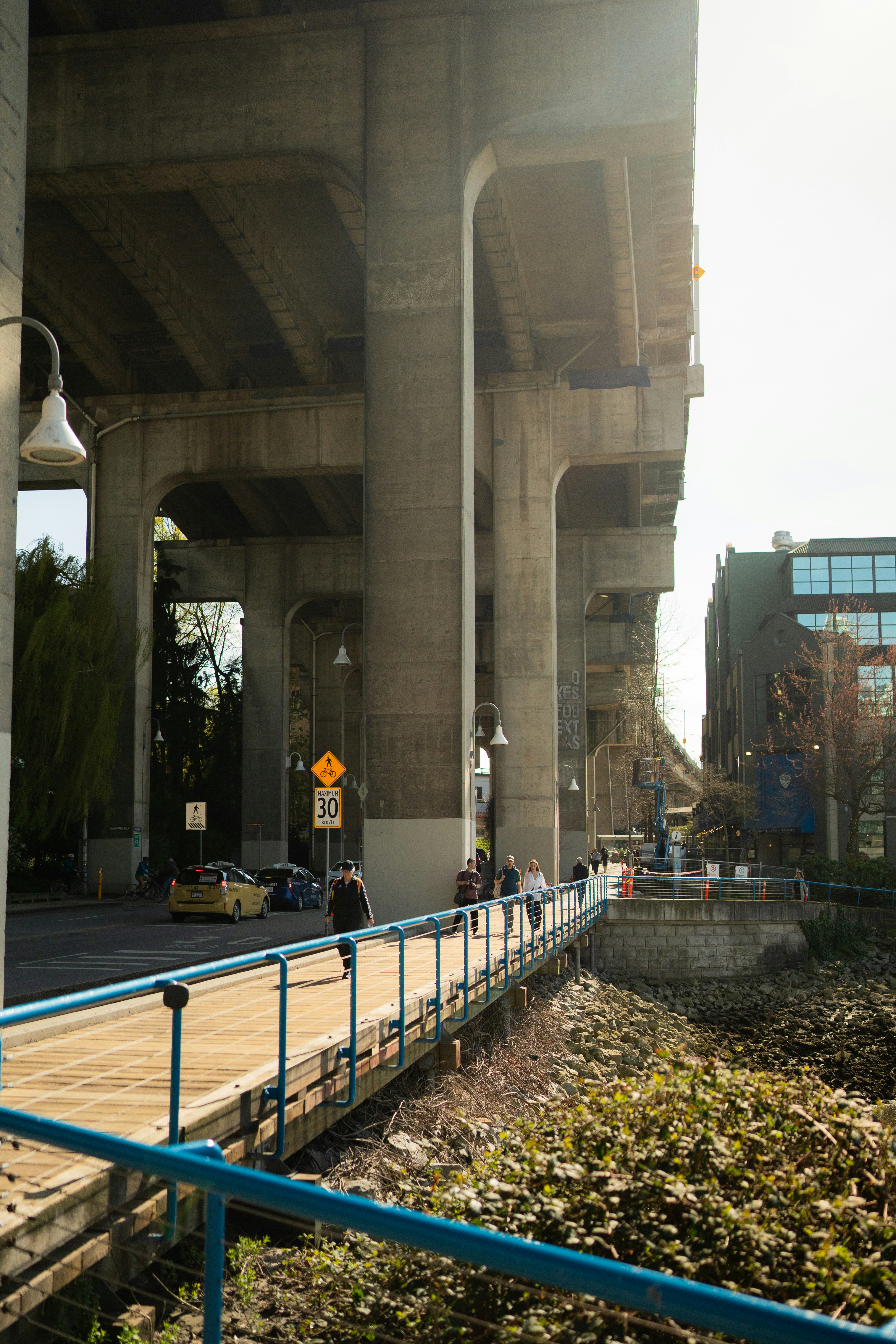 Pedestrians stroll along a wooden walkway beneath a concrete overpass, framed by urban architecture and greenery.