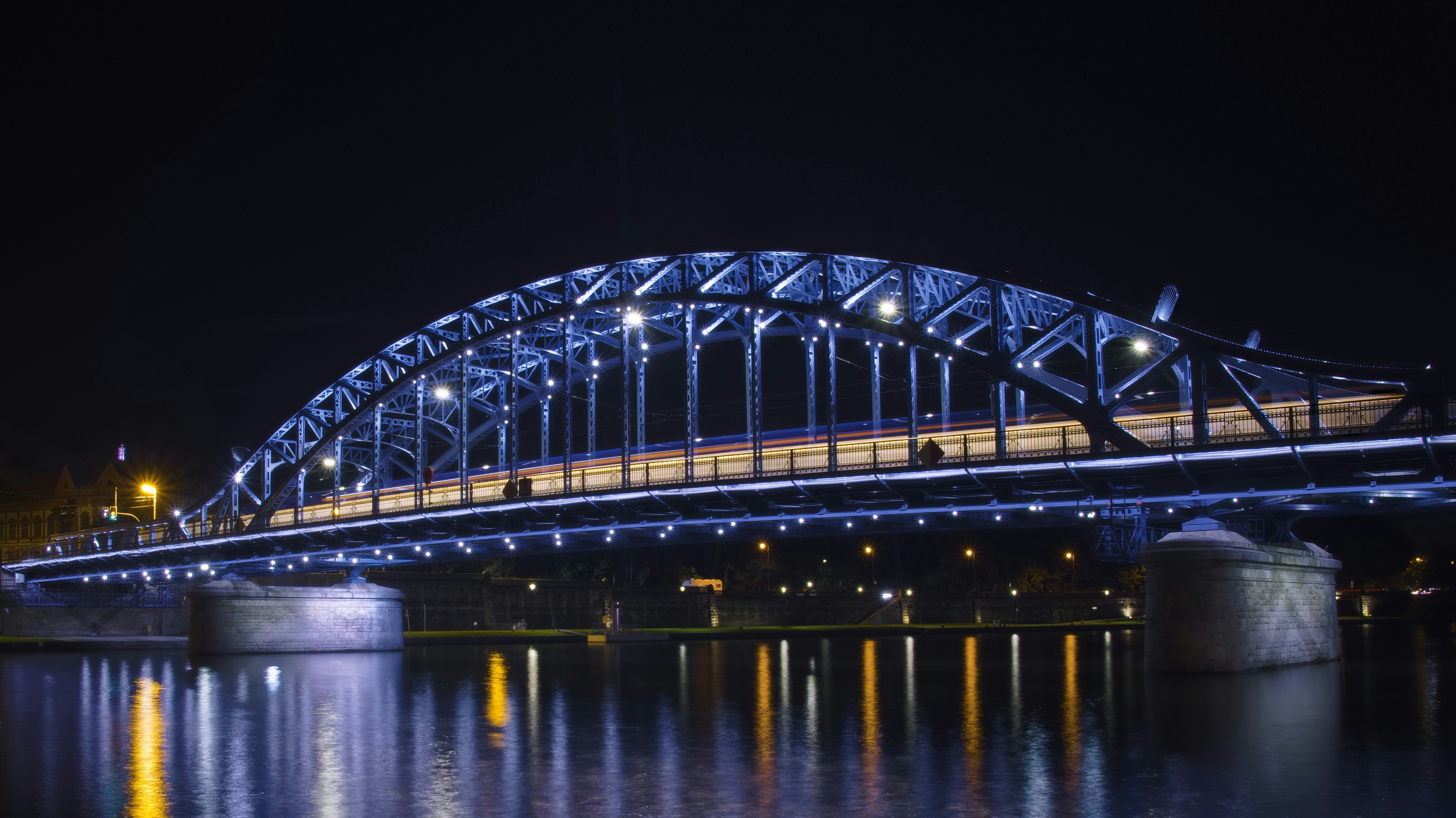 A striking long-exposure night shot of a steel bridge bathed in brilliant blue light, capturing a glowing train mid-transit over the water. The reflection dances on the calm surface below, while the sharp geometry of the bridge contrasts against the black void of the sky. A seamless mix of urban motion and stillness.