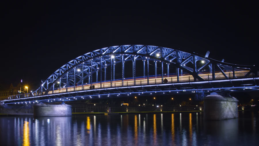 Blue-lit arch bridge spans a calm river at night, with reflections shimmering on the water below. Warm lights line the roadway against a dark sky.