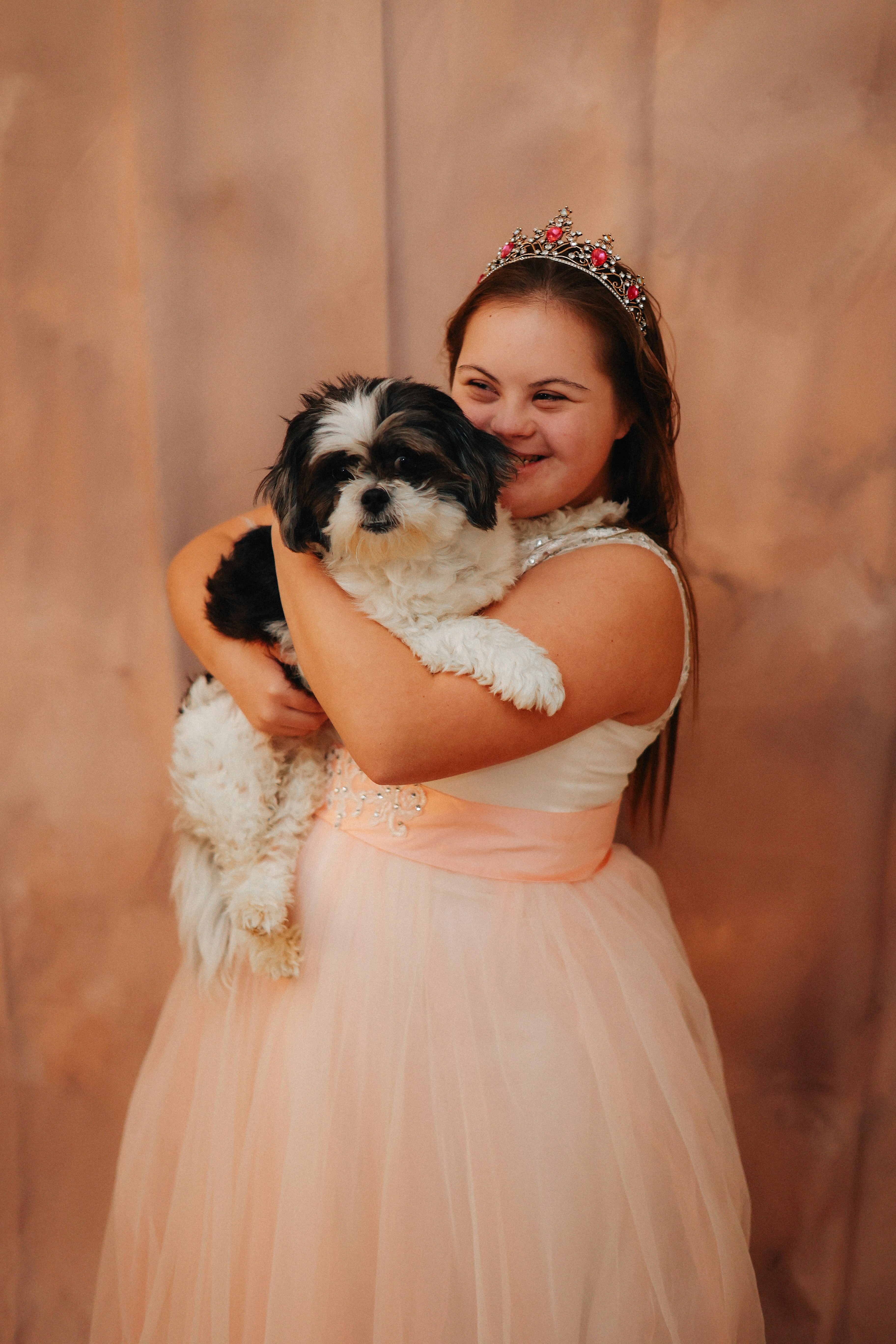 Girl in a pink dress joyfully holding a small dog, showcasing a heartwarming connection. The soft background complements the playful scene.
