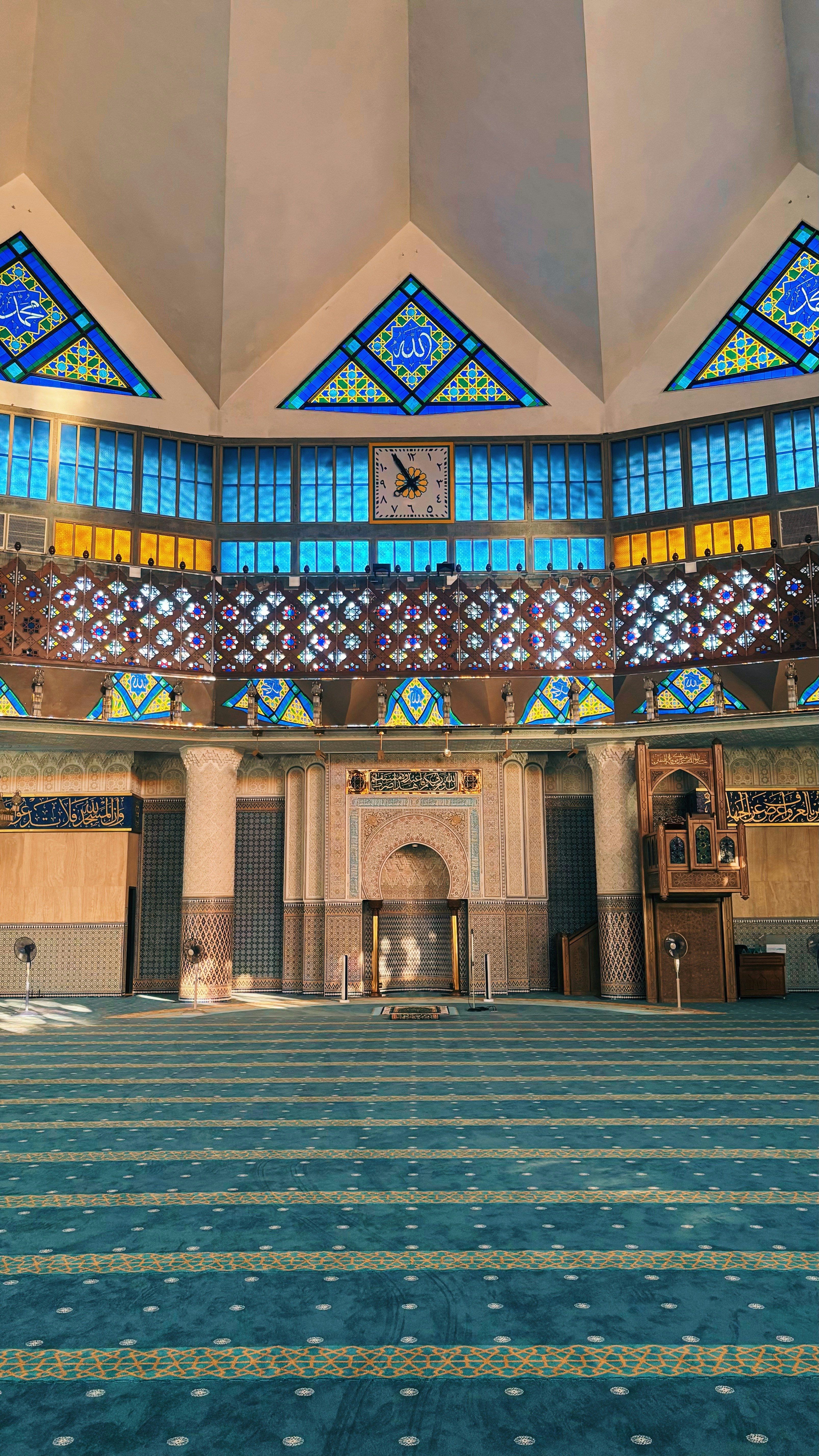 Interior view of a mosque showcasing intricate geometric patterns and vibrant stained glass windows. The serene atmosphere is enhanced by the expansive carpet and detailed architectural elements.