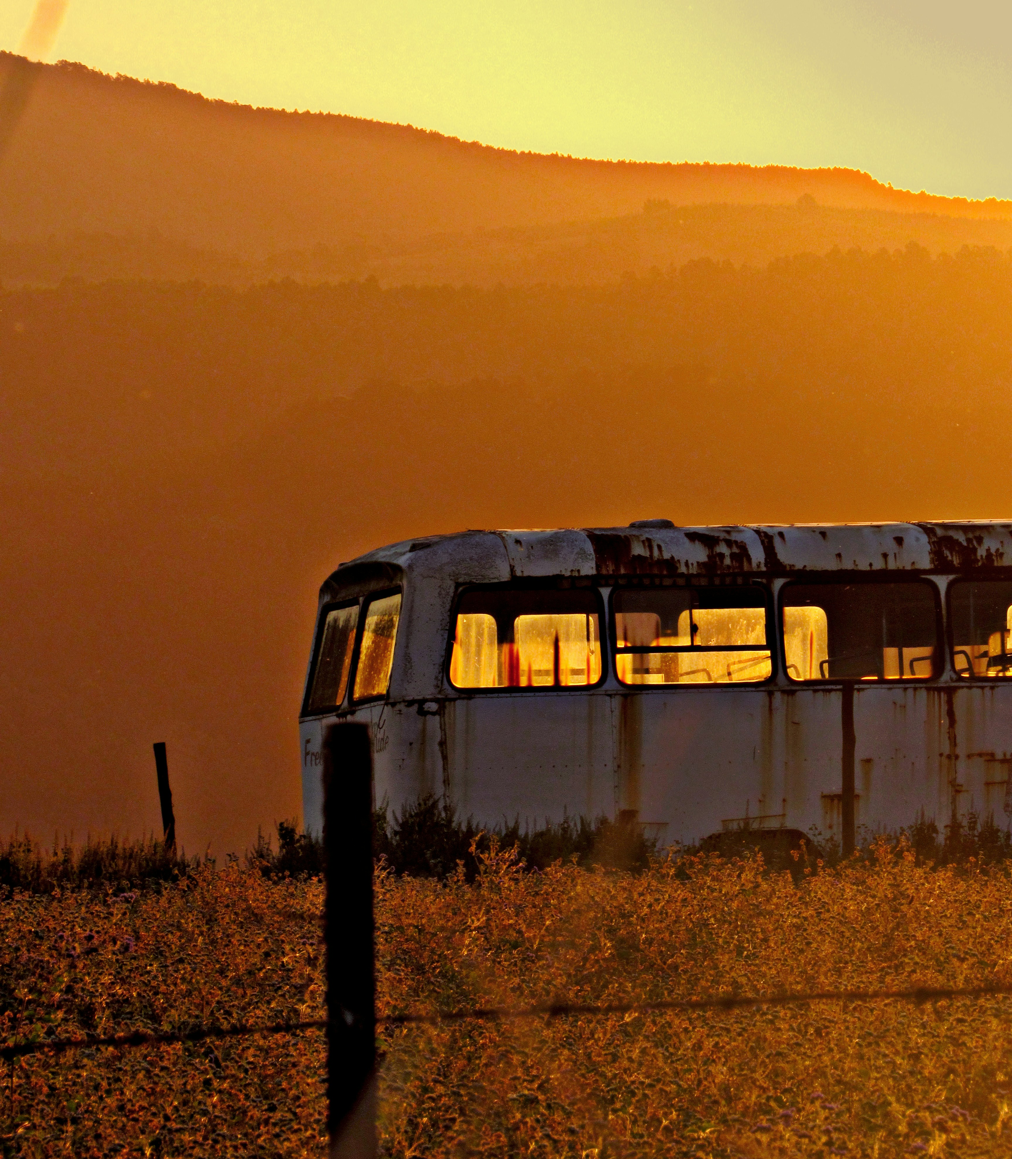 Old rusted bus sits in a sunlit field during golden hour, its windows glowing with warm interior light. The image emphasizes abandonment and quiet landscape at sunset.
