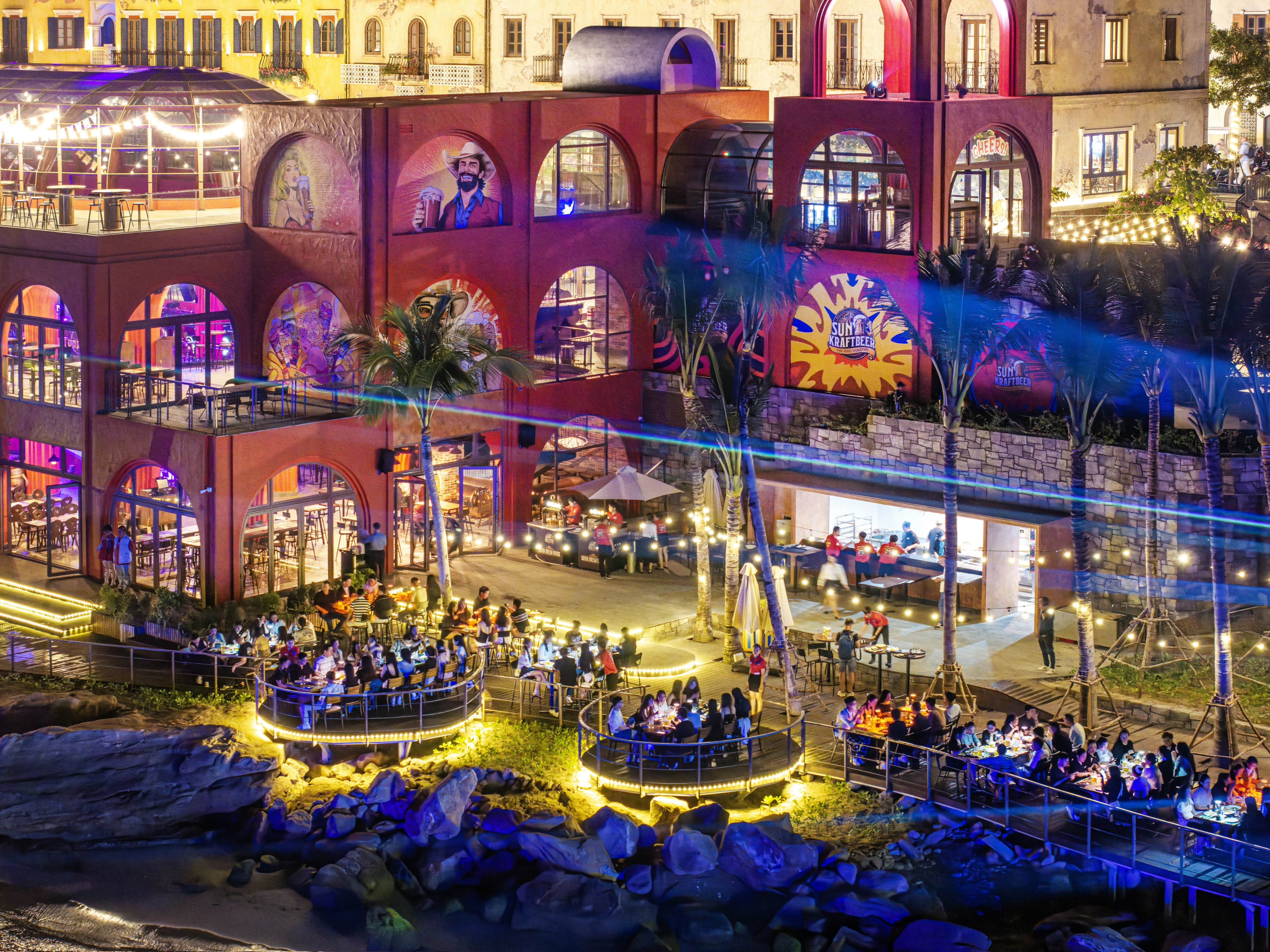 Lively outdoor dining scene illuminated by colorful lights, featuring a bustling crowd enjoying a night out at a coastal venue.