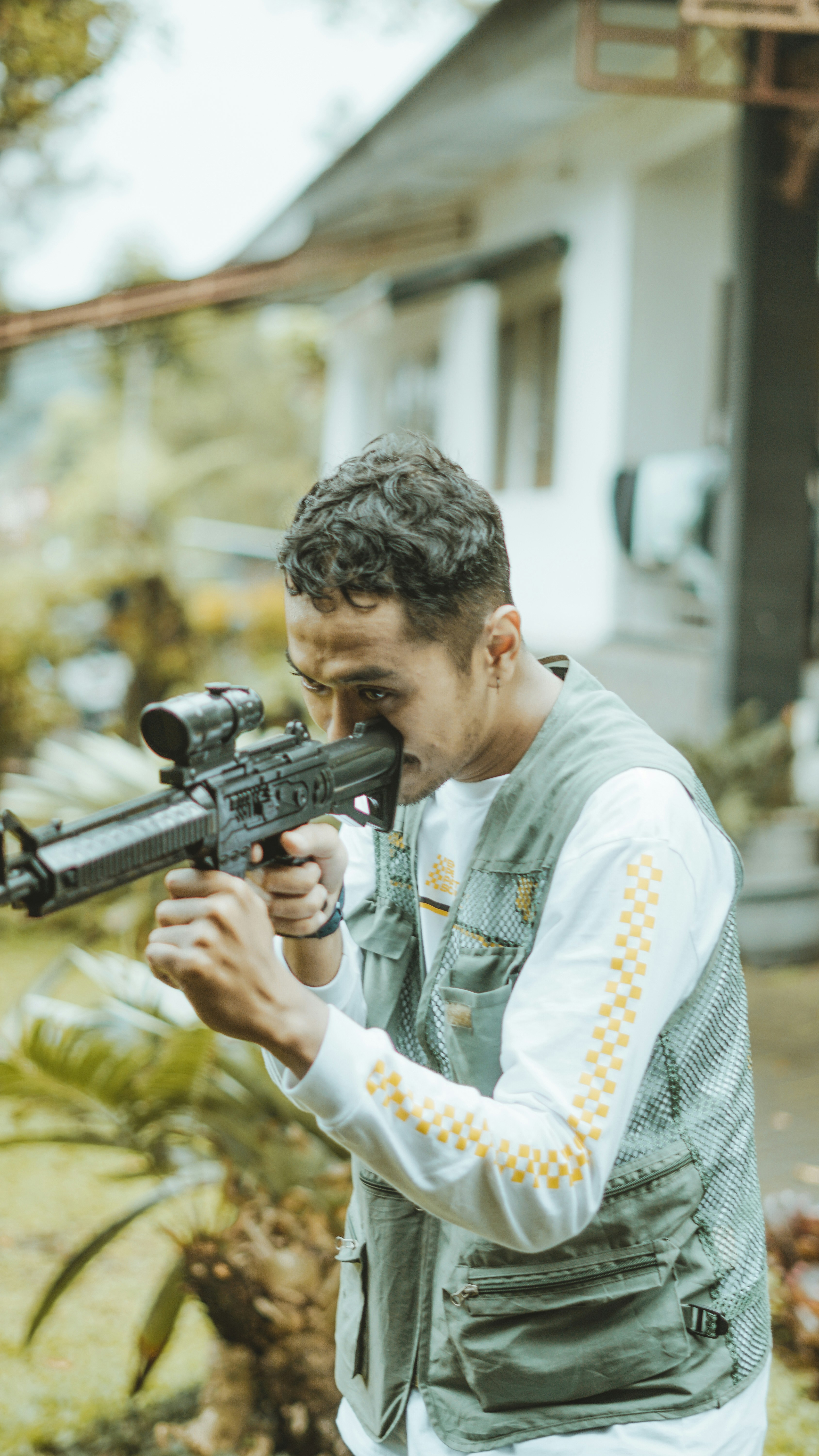 A young man aiming a rifle with intense concentration, set against a blurred natural backdrop. The scene conveys a sense of action and determination.