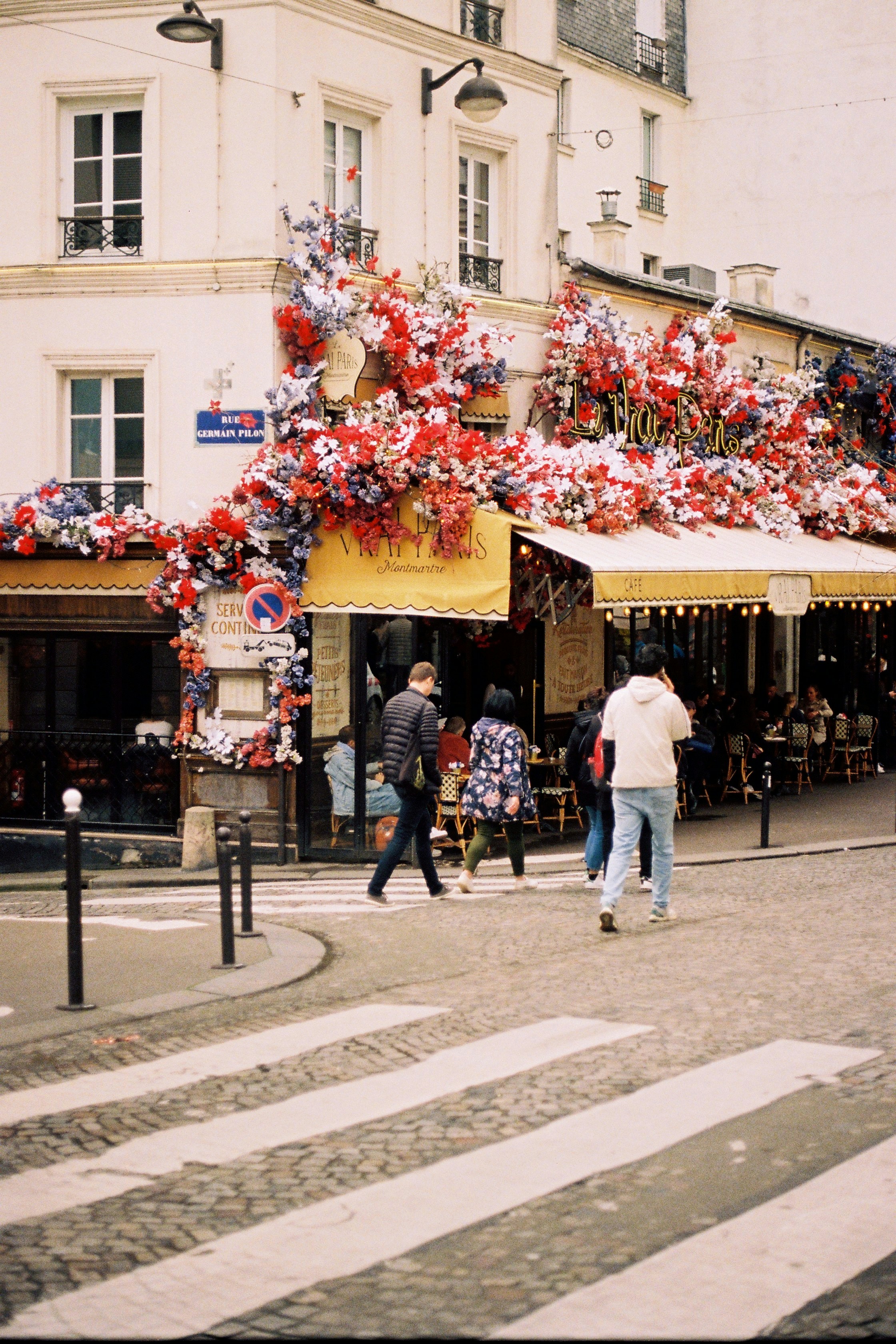 A charming café adorned with vibrant floral decorations, bustling with pedestrians on a cobblestone street in Paris.