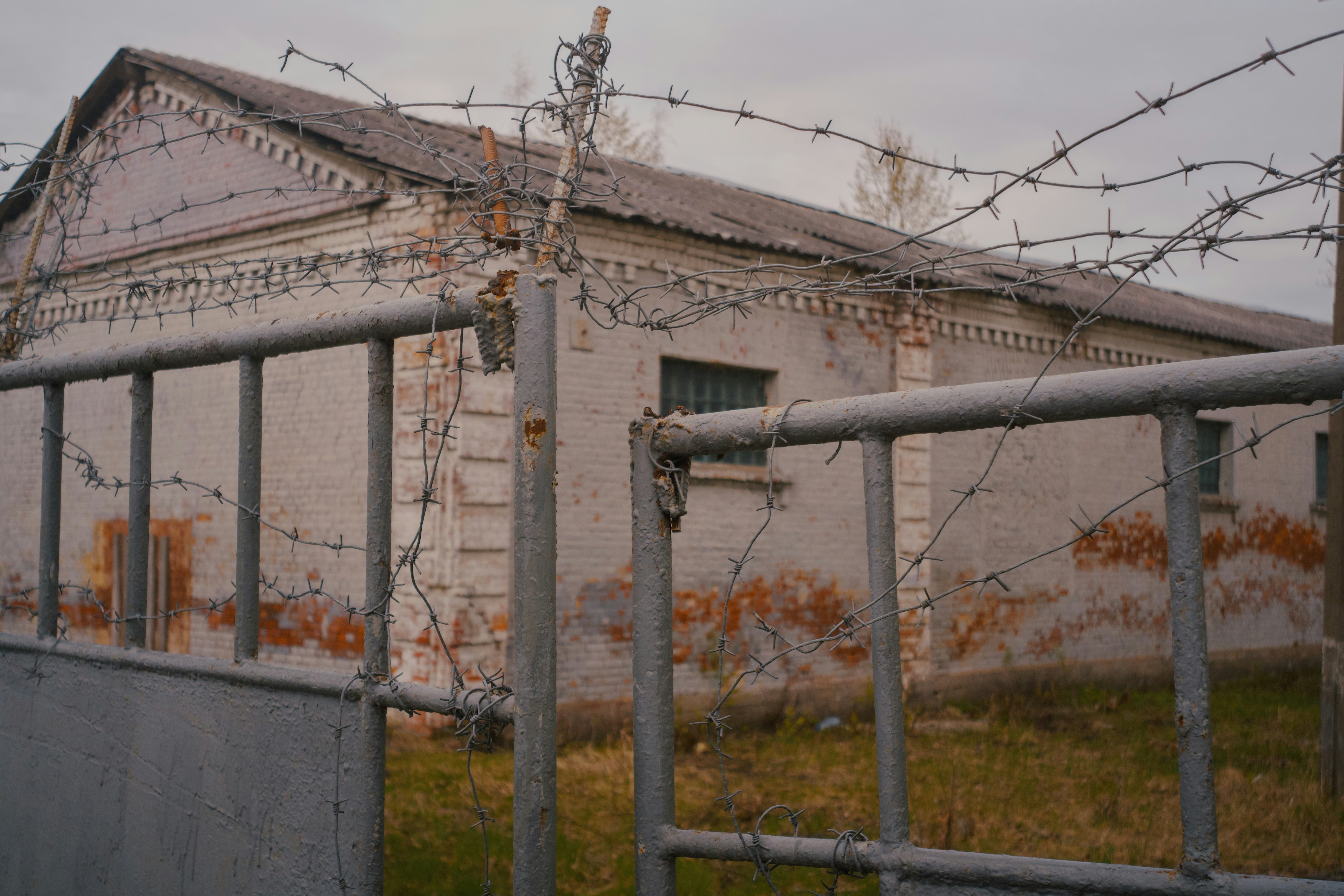 A decrepit building is behind a gate and barbed wire. photo – Free ...