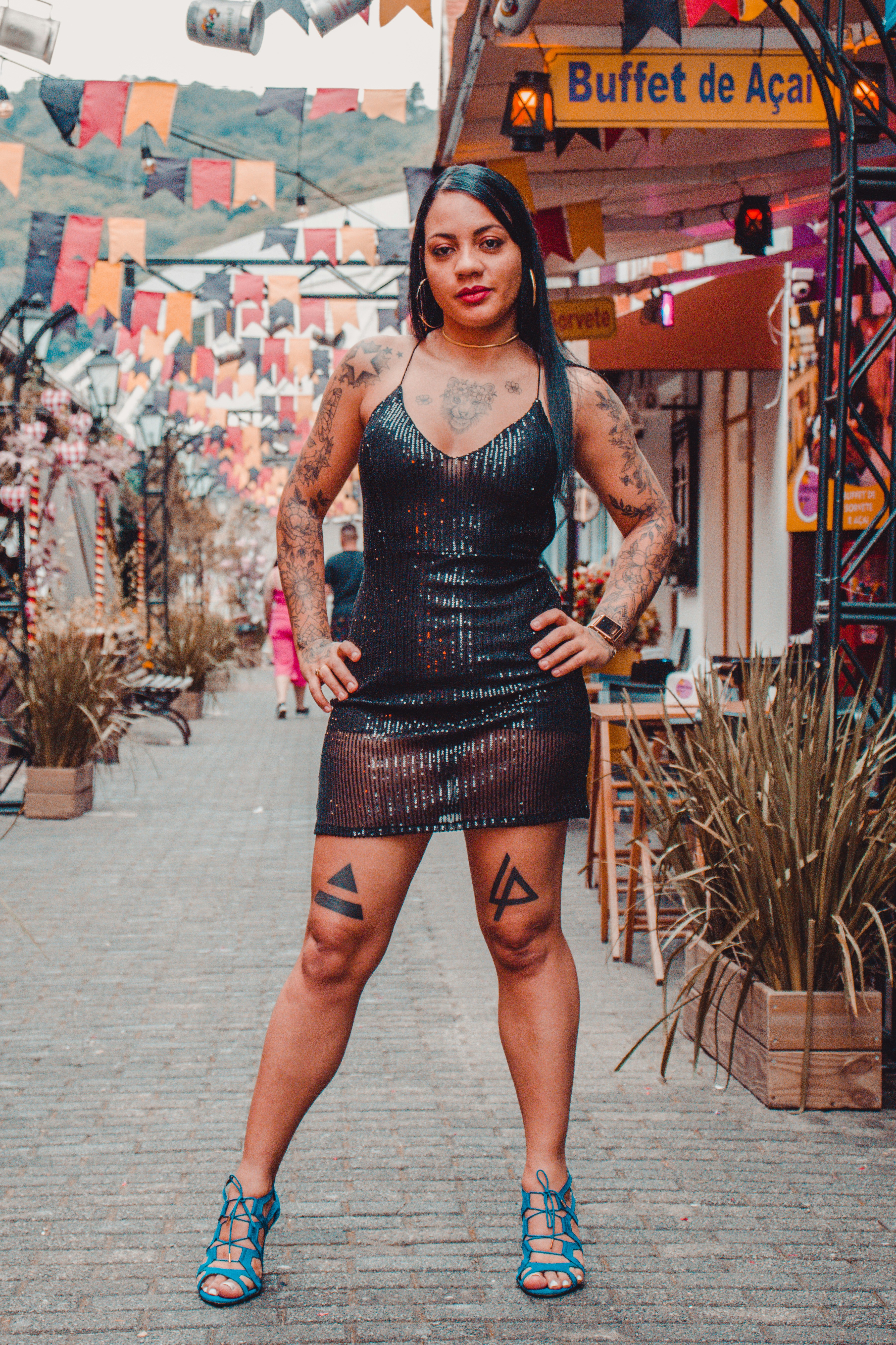 A confident woman stands in a lively street adorned with colorful flags and decorations, showcasing her tattoos and stylish outfit.