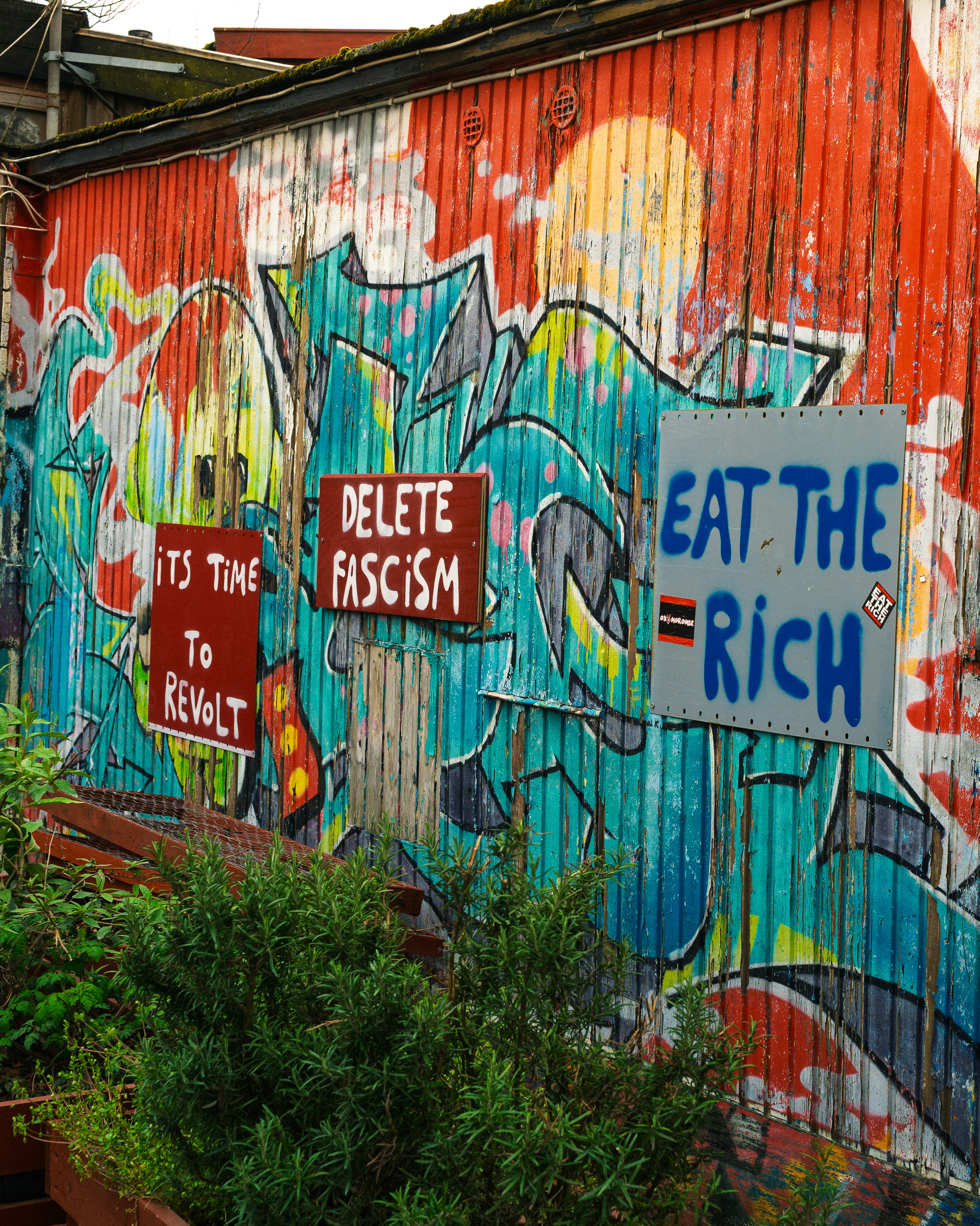 Colorful graffiti painting on a corrugated wall features protest signs reading 'DELETE FASCISM' and 'EAT THE RICH'.