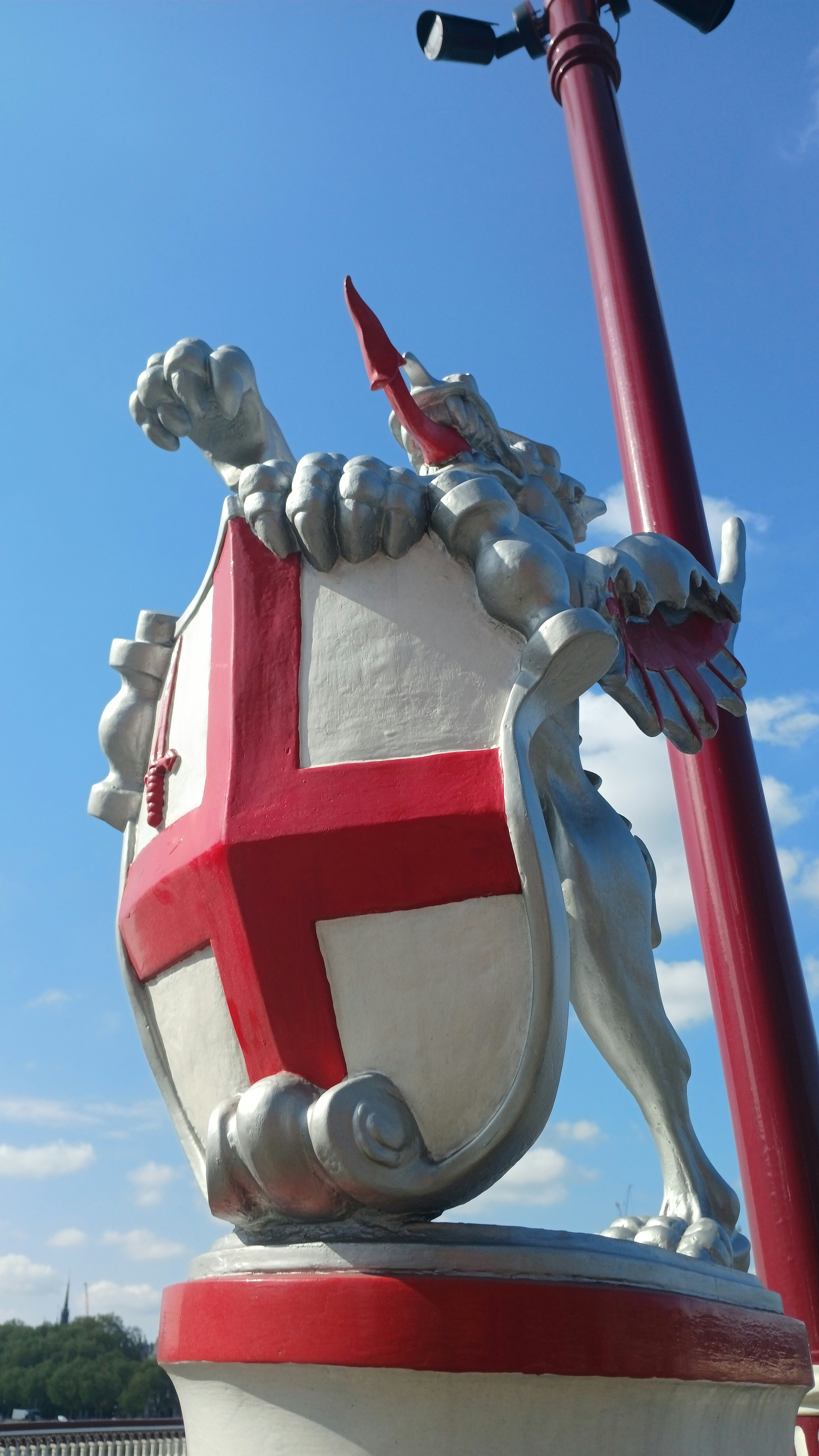 A silver lion sculpture adorned with a red cross shield, standing proudly atop a pedestal against a bright blue sky.