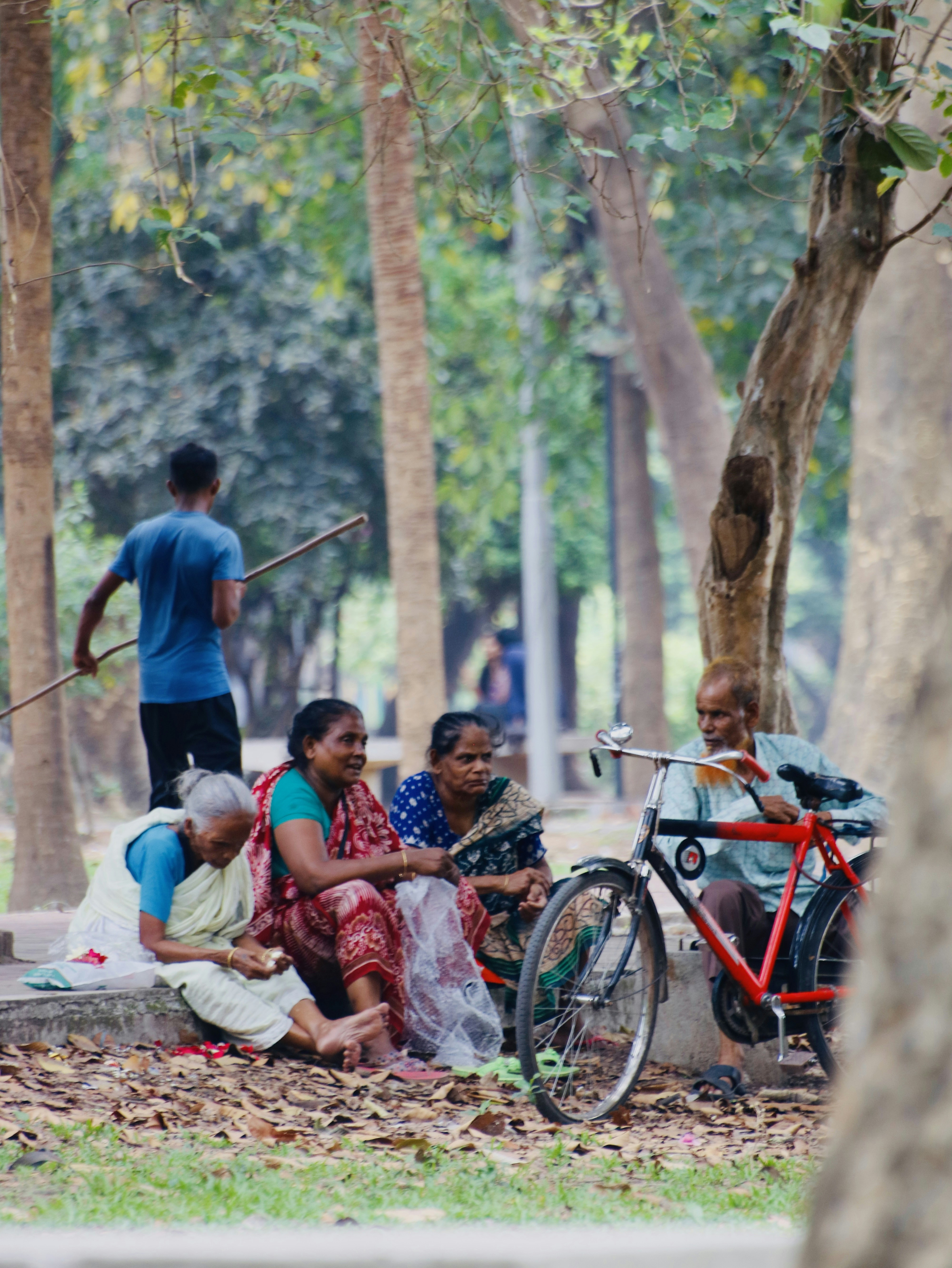 Village gathering under trees: elders sharing stories with a passing cyclist and a red bicycle