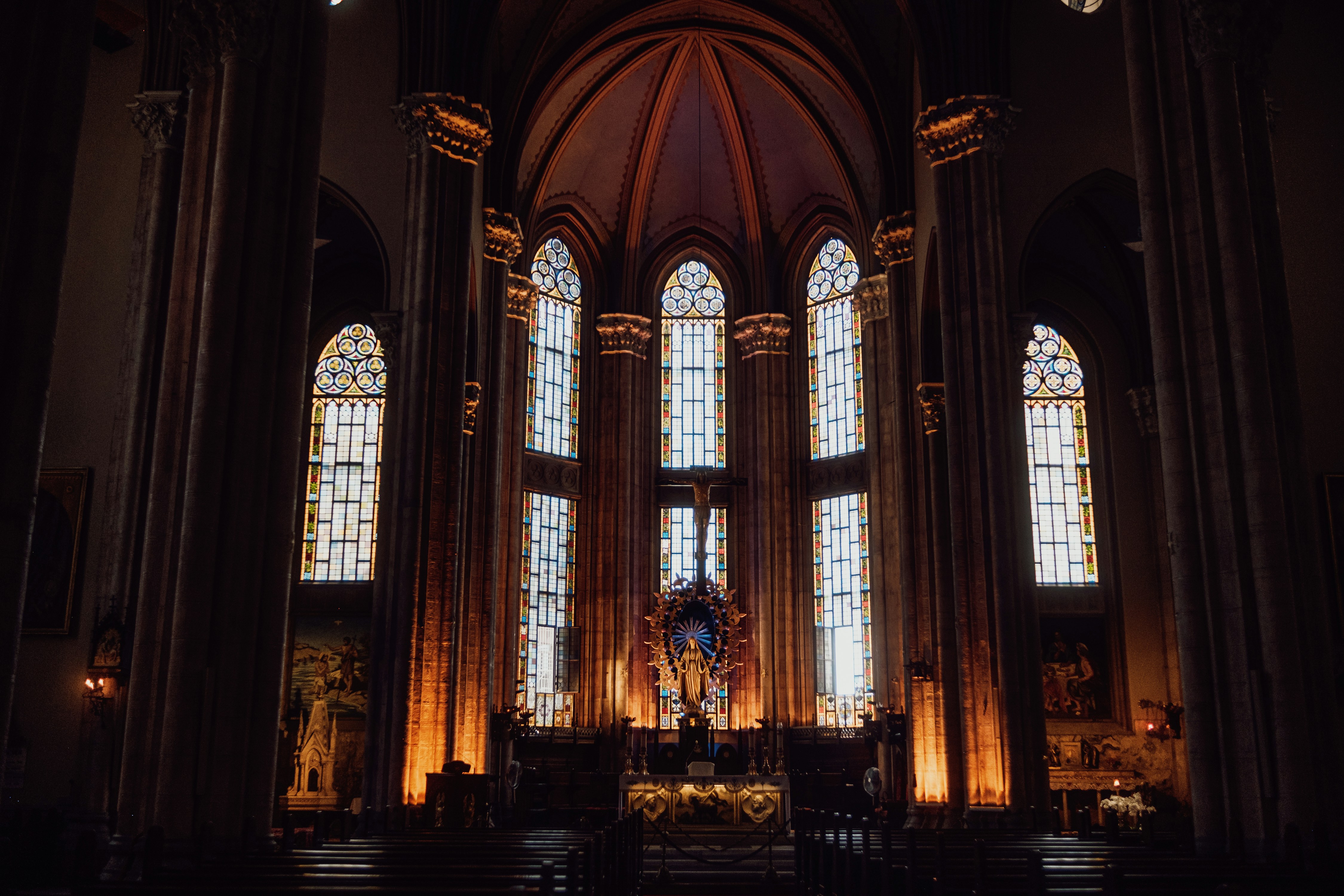 Intricate stained glass windows illuminate the interior of a grand church, highlighting the altar and architectural details. Warm light enhances the serene atmosphere.