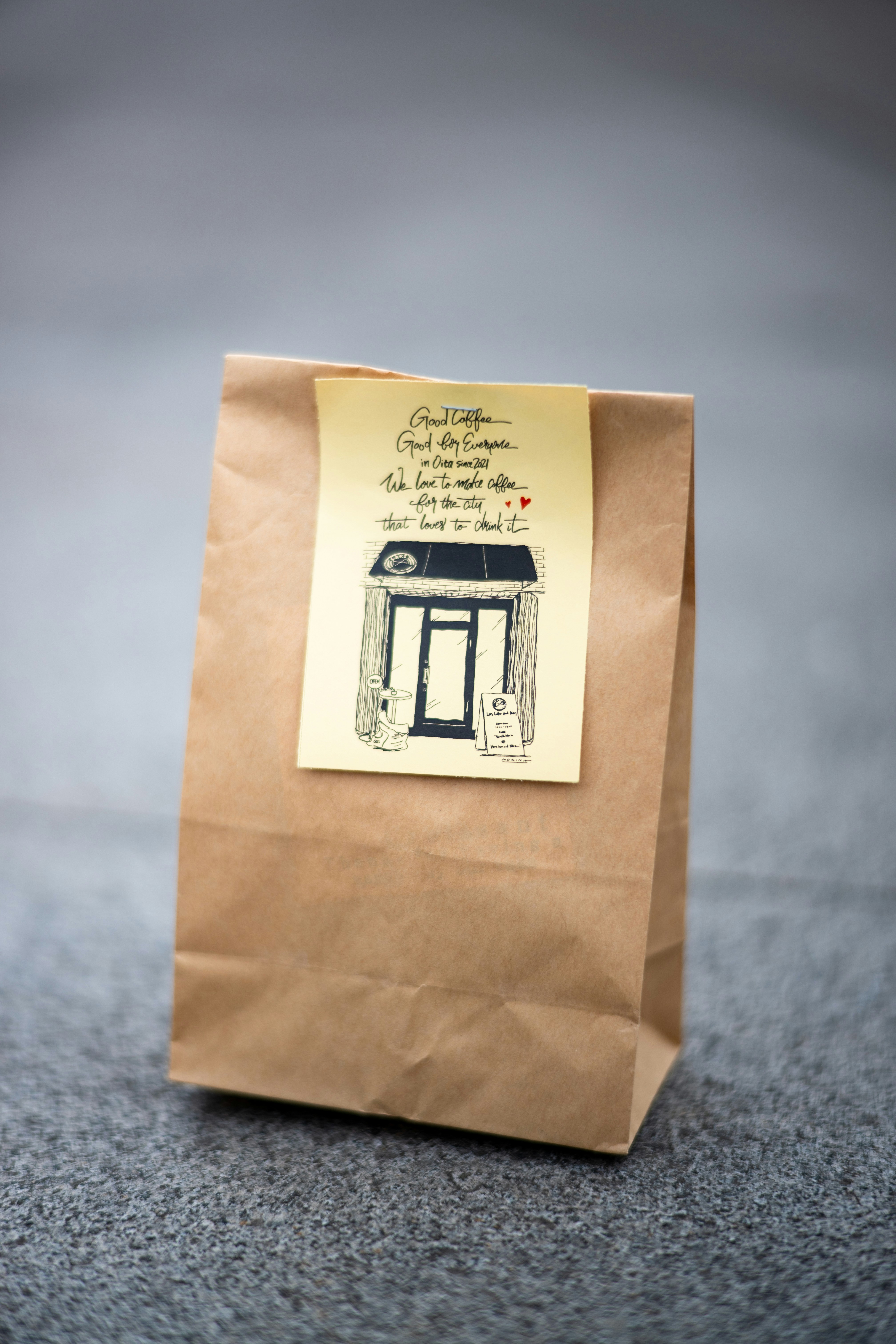 Brown paper bag featuring a handwritten note with a charming illustration of a coffee shop entrance. The note expresses a friendly message to customers.
