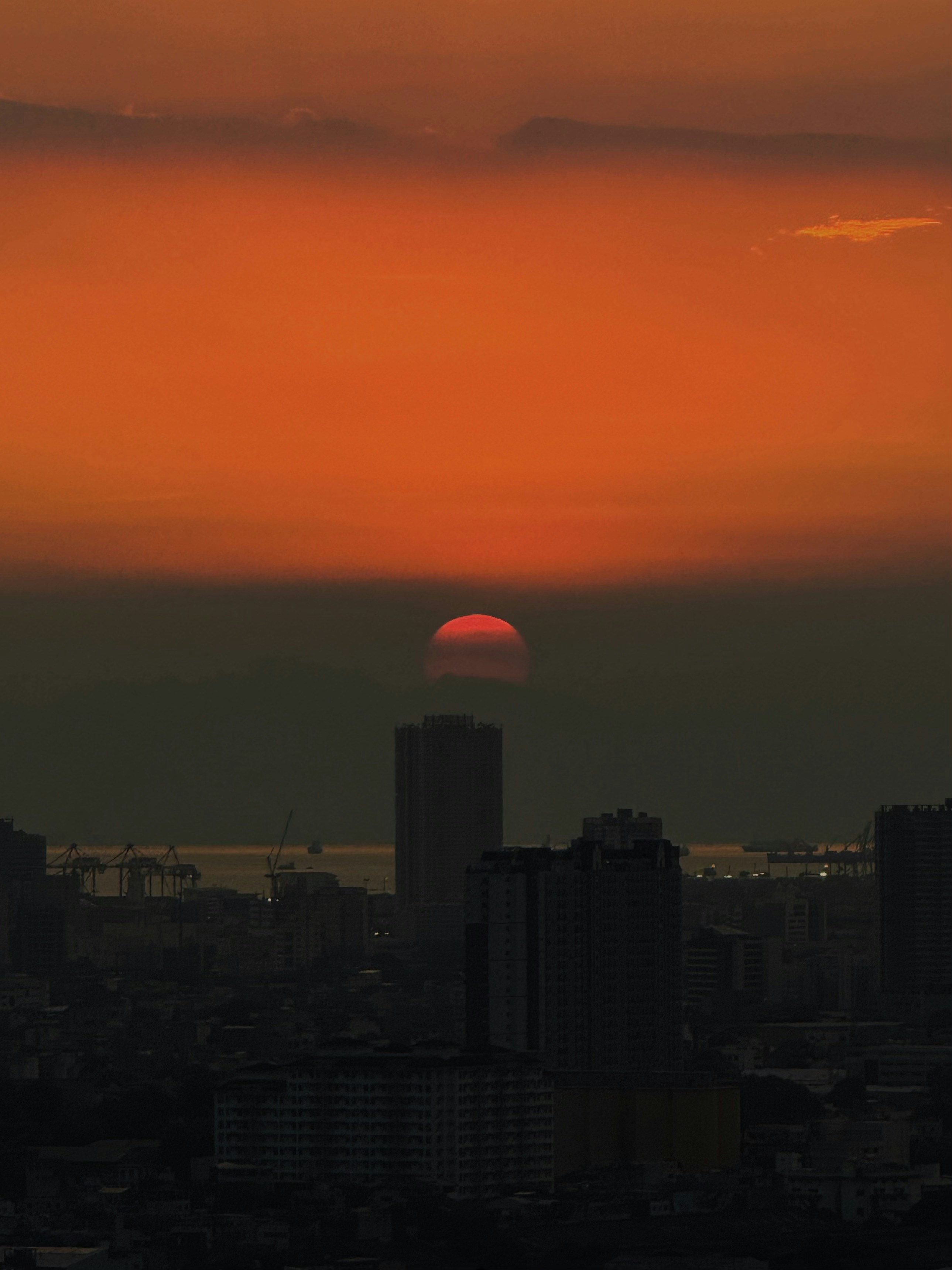 Orange-hued sunset casting a warm glow over a city skyline, with the sun partially hidden behind a building. The scene captures the tranquil transition from day to night.