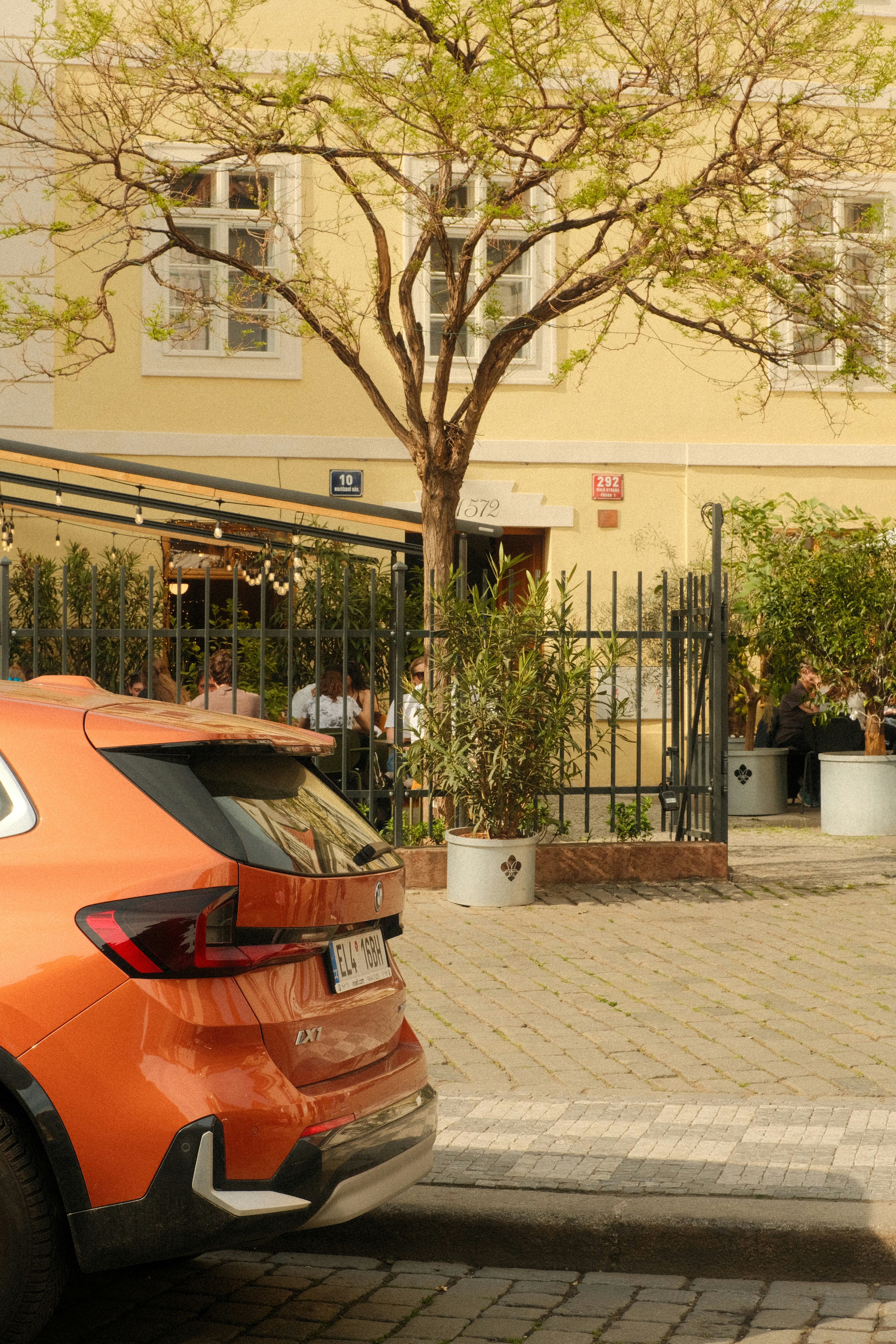 Customers enjoy their time at a lively outdoor cafe nestled against yellow walls. An orange car is parked nearby, while greenery adds freshness to the setting. BMW X1.