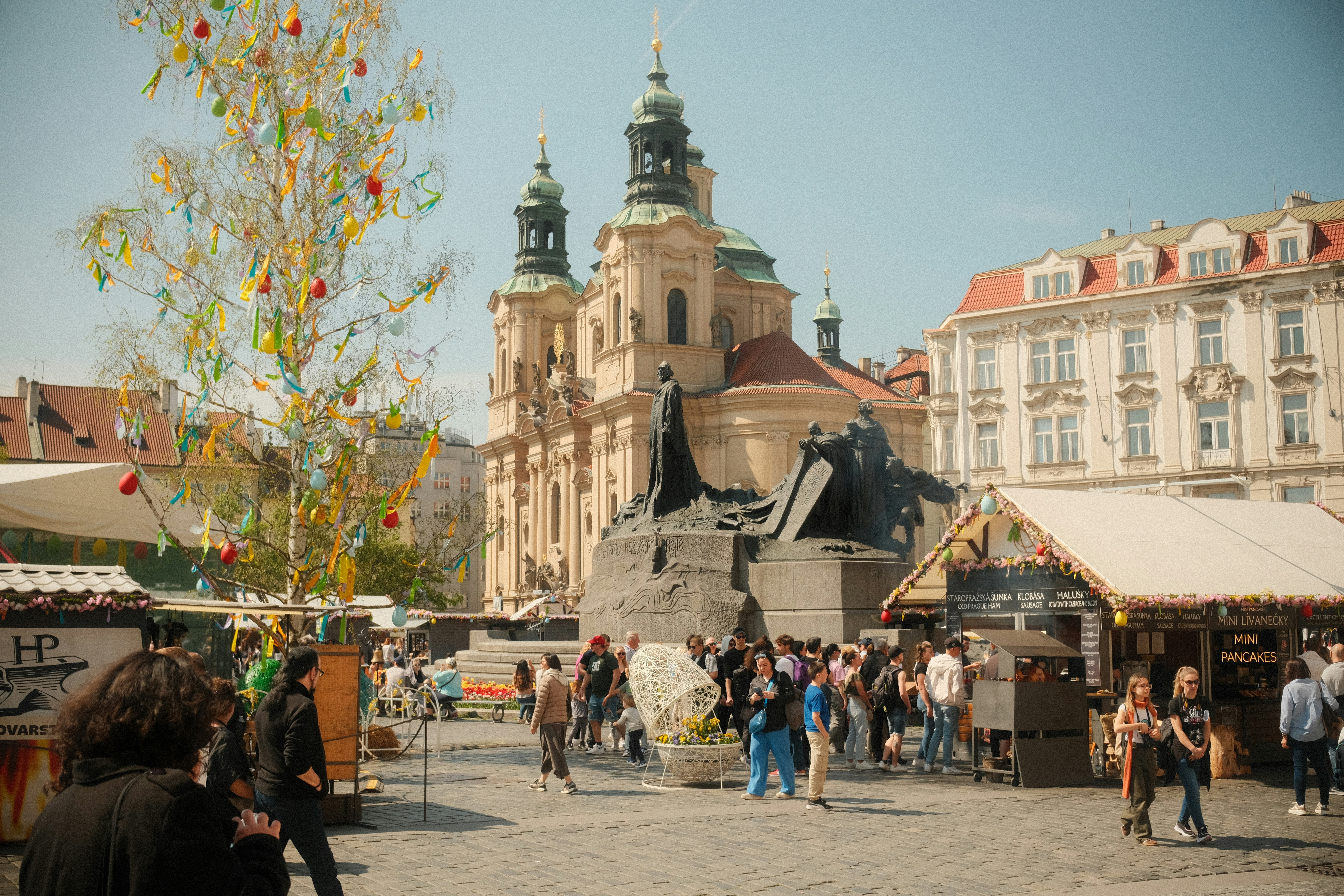 Crowd gathers at historic square with market stalls and colorful tree decorations in Prague on a sunny day. People stroll through a vibrant square in Prague, surrounded by festive market stalls and a beautifully decorated tree, all under the bright sunlight.