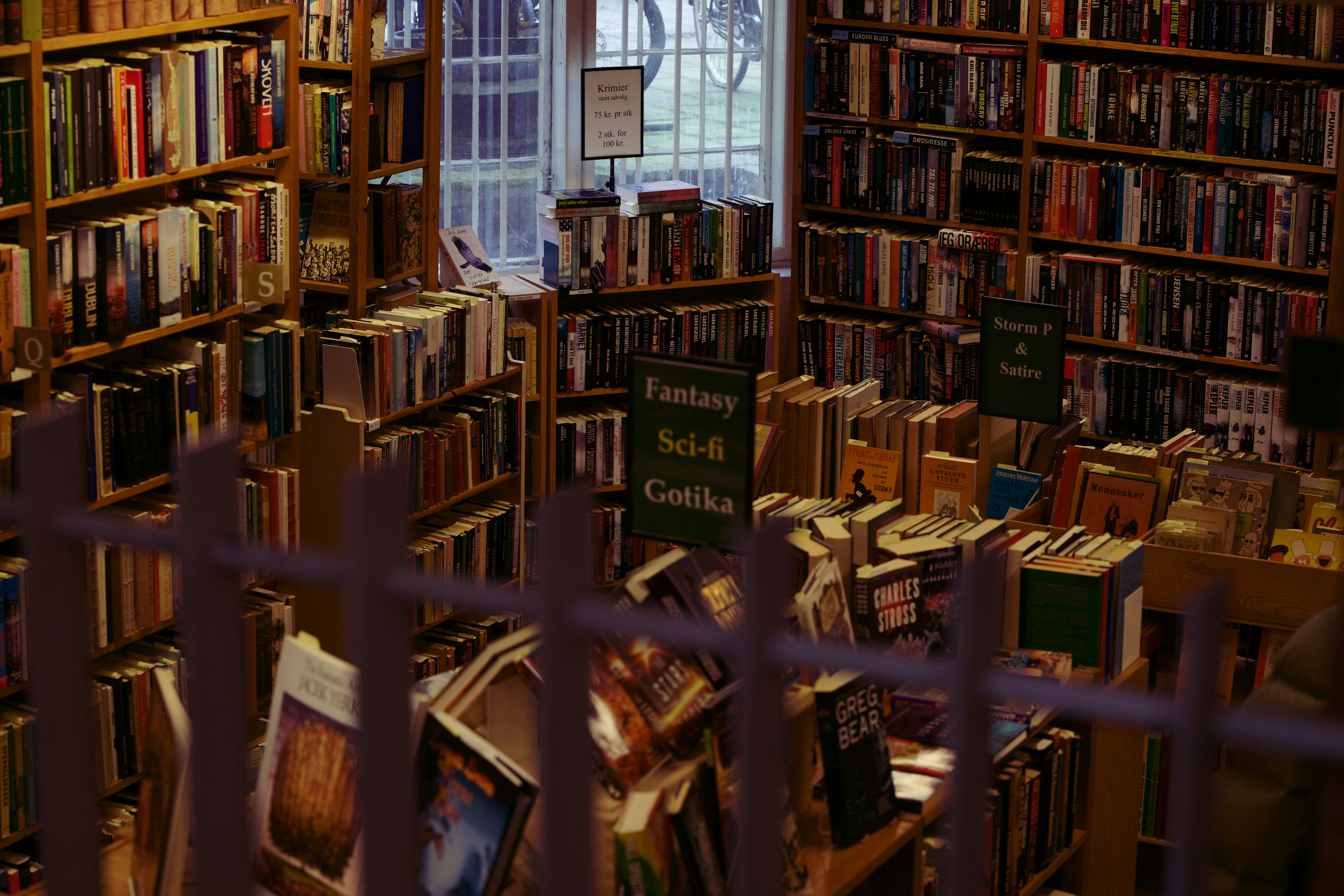 A cozy bookstore filled with shelves of books, featuring signs for various genres like Fantasy and Sci-fi. Warm lighting creates an inviting atmosphere.