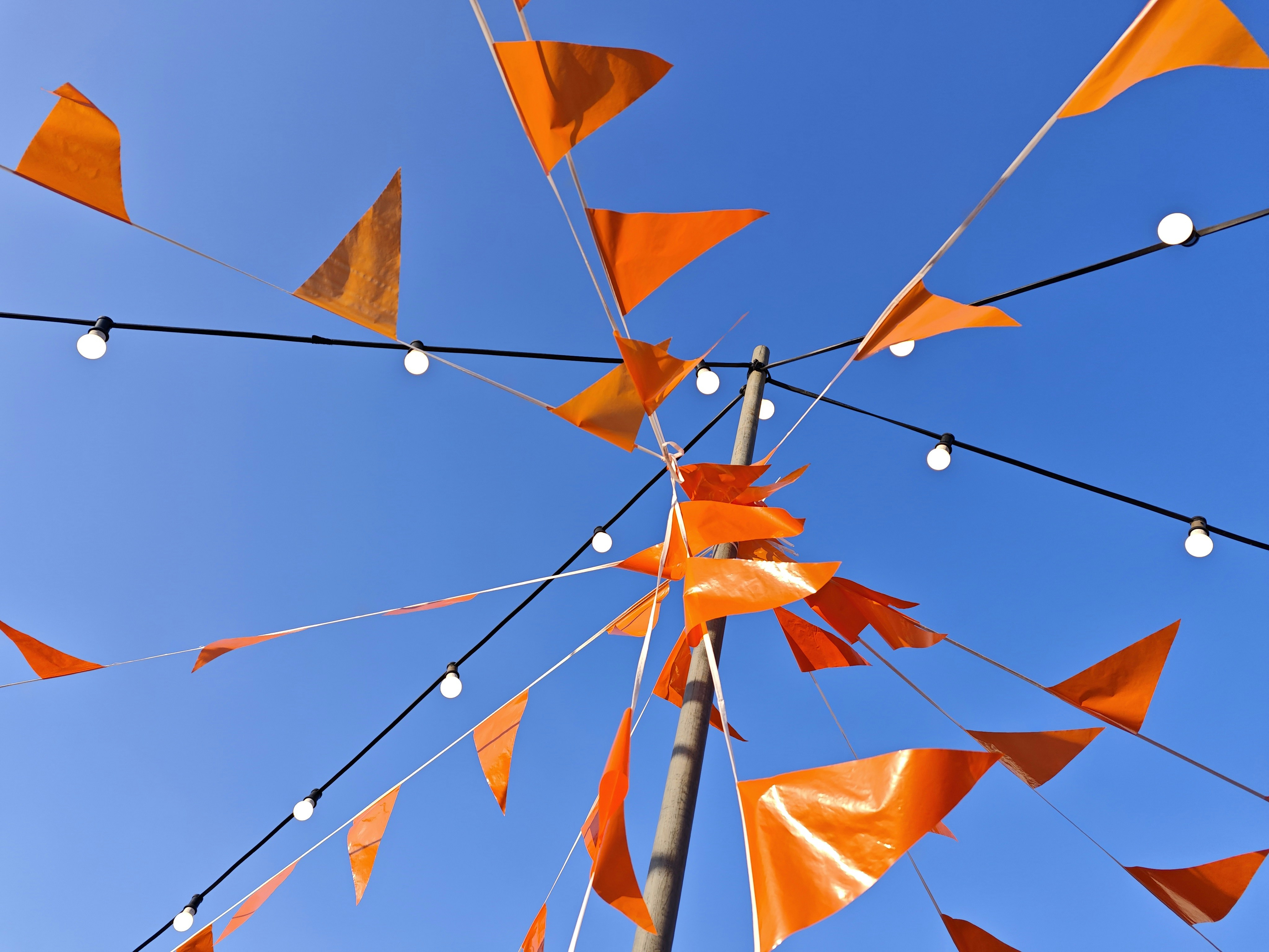 Vibrant orange flags fluttering against a clear blue sky, interspersed with glowing string lights, create a festive atmosphere.