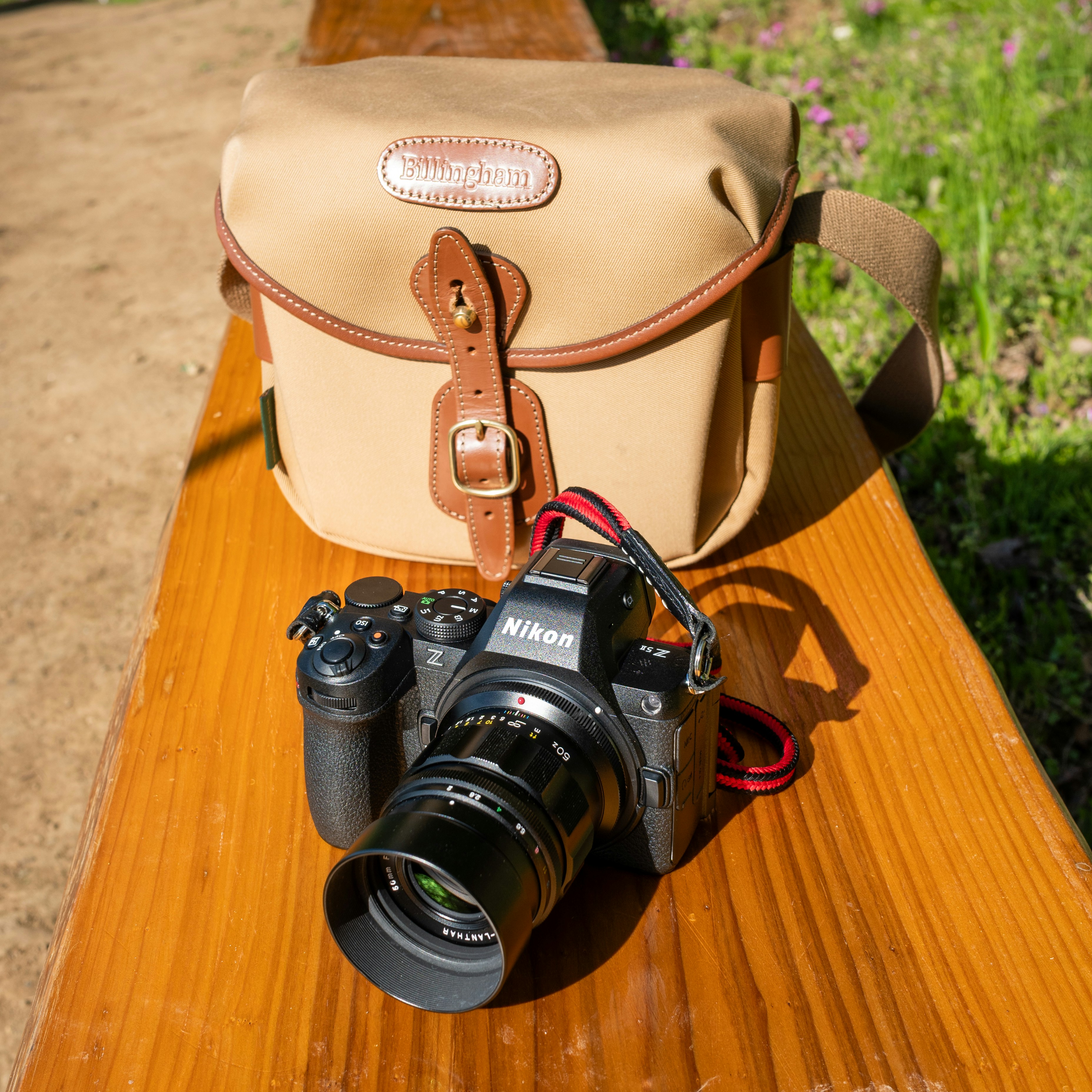 Photograph shows a Nikon camera with a prominent lens resting on a polished wooden bench beside a tan leather bag in bright outdoor light.