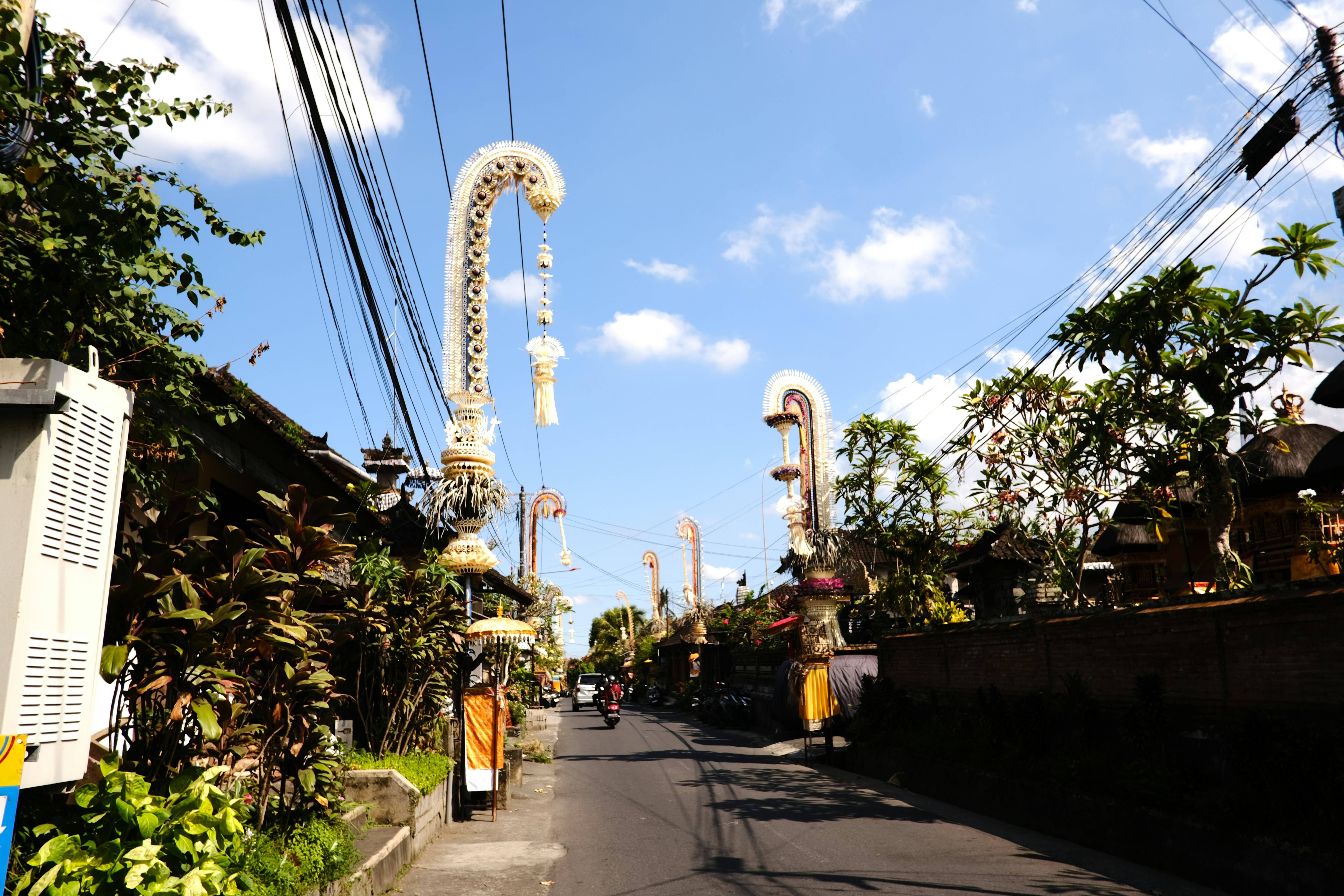 Colorful ceremonial poles line a Bali street, framed by lush greenery and a clear blue sky. The scene captures the vibrant cultural essence of the area.