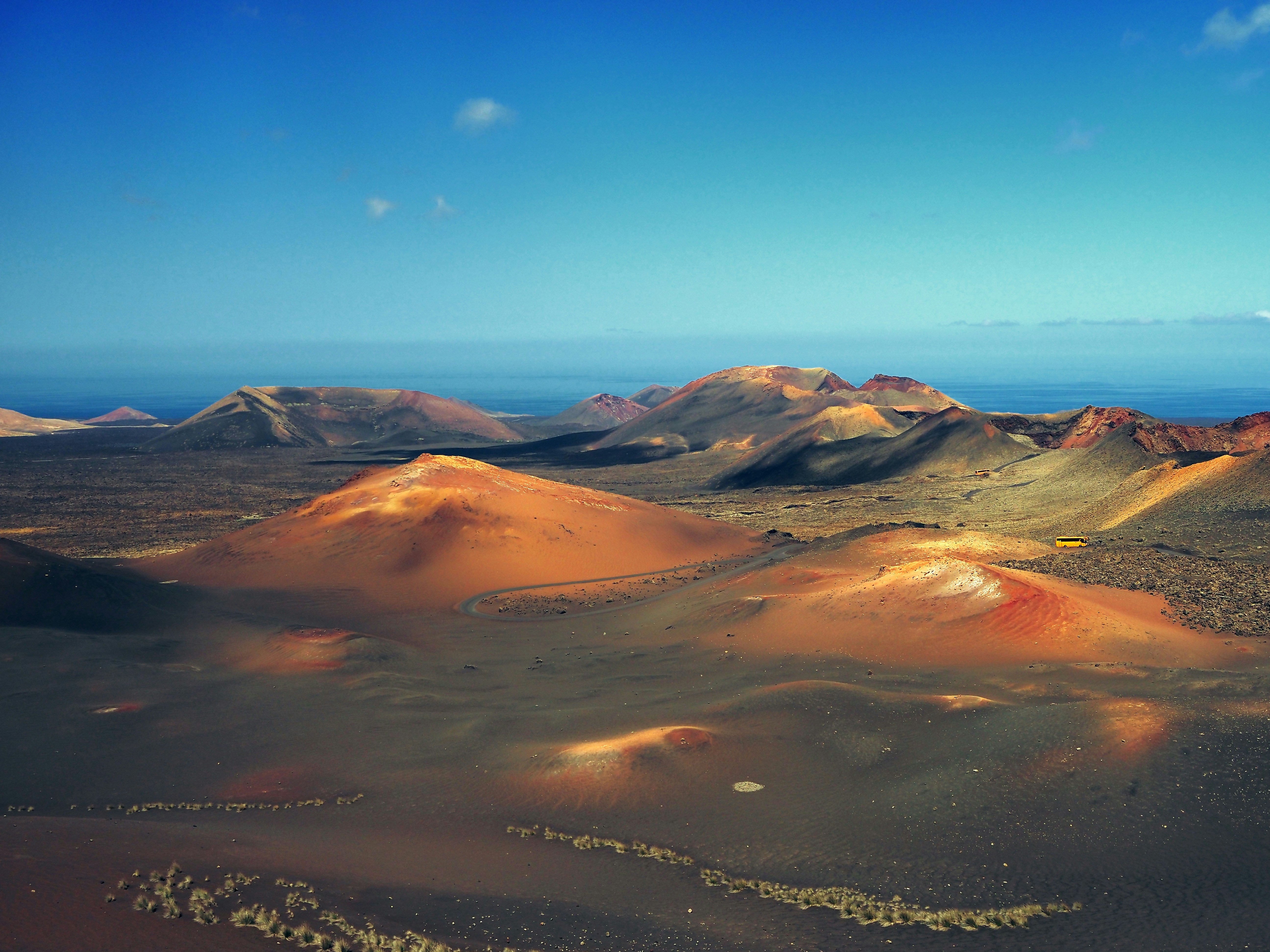 Volcanic dunes in warm red-orange tones stretch across the foreground in this landscape photograph, under a deep blue sky. The stark color contrast and textured surfaces emphasize the arid volcanic terrain.
