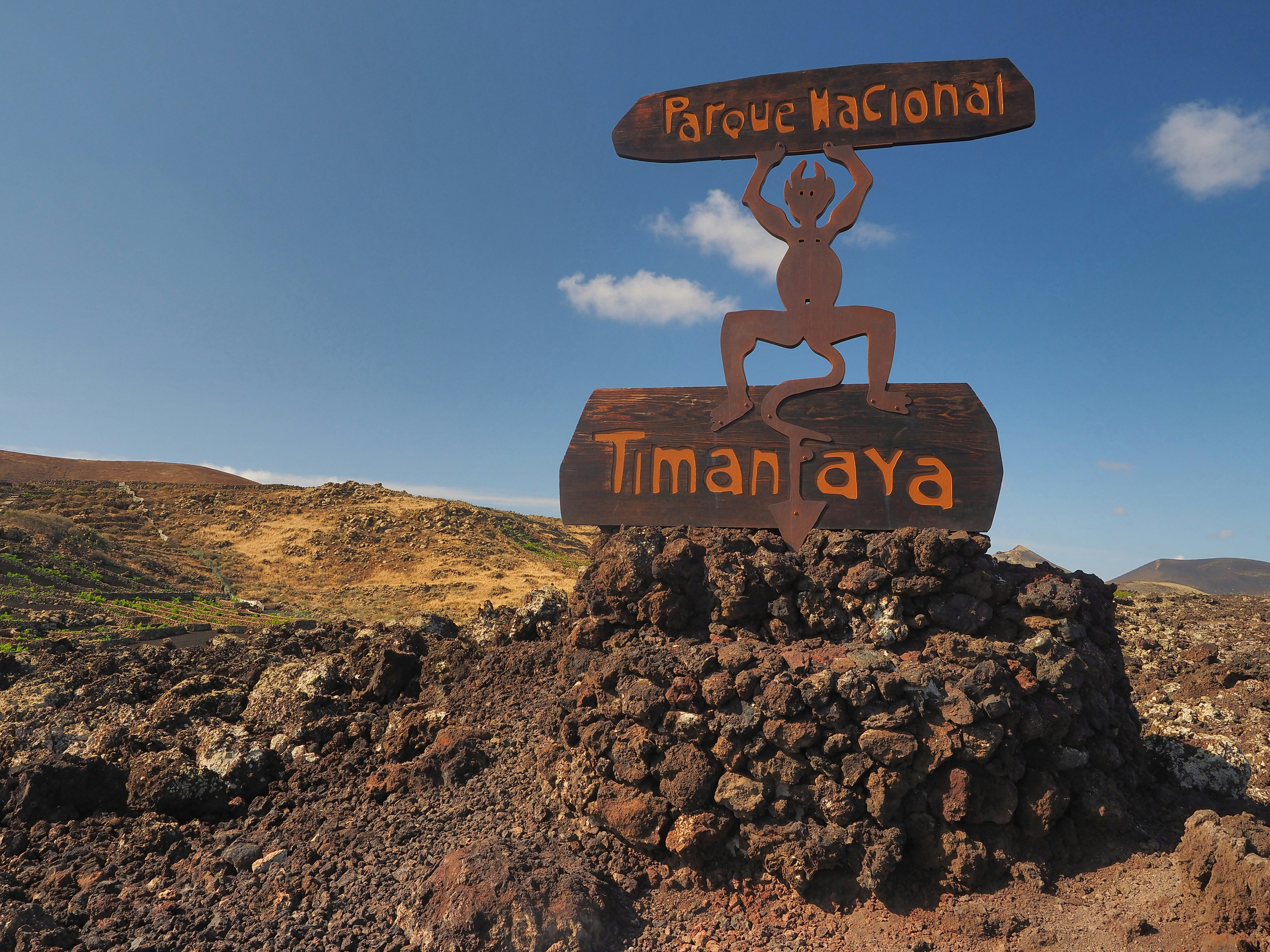 A carved wooden sign reading 'Parque Nacional Timanfaya' sits on a lava-rock pedestal, set against a stark volcanic landscape.