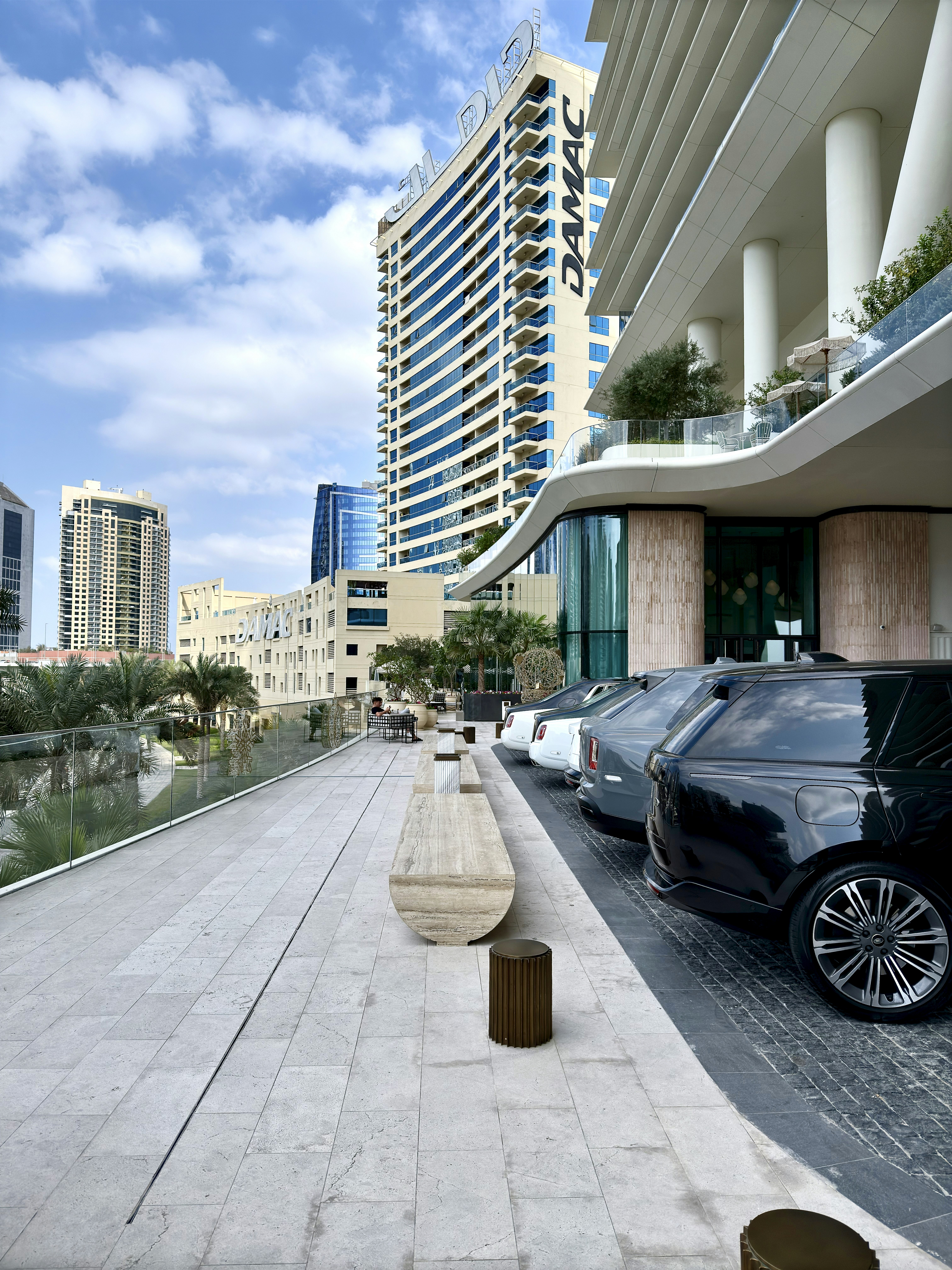 Chic Urban Retreat: A stylish outdoor space with modern seating and luxury cars parked alongside a sleek high-rise, offering a glimpse of upscale city living. The towering buildings and palm trees under a partly cloudy sky create a perfect blend of urban sophistication and tropical charm. Ideal for those who love modern architecture and luxury lifestyles.