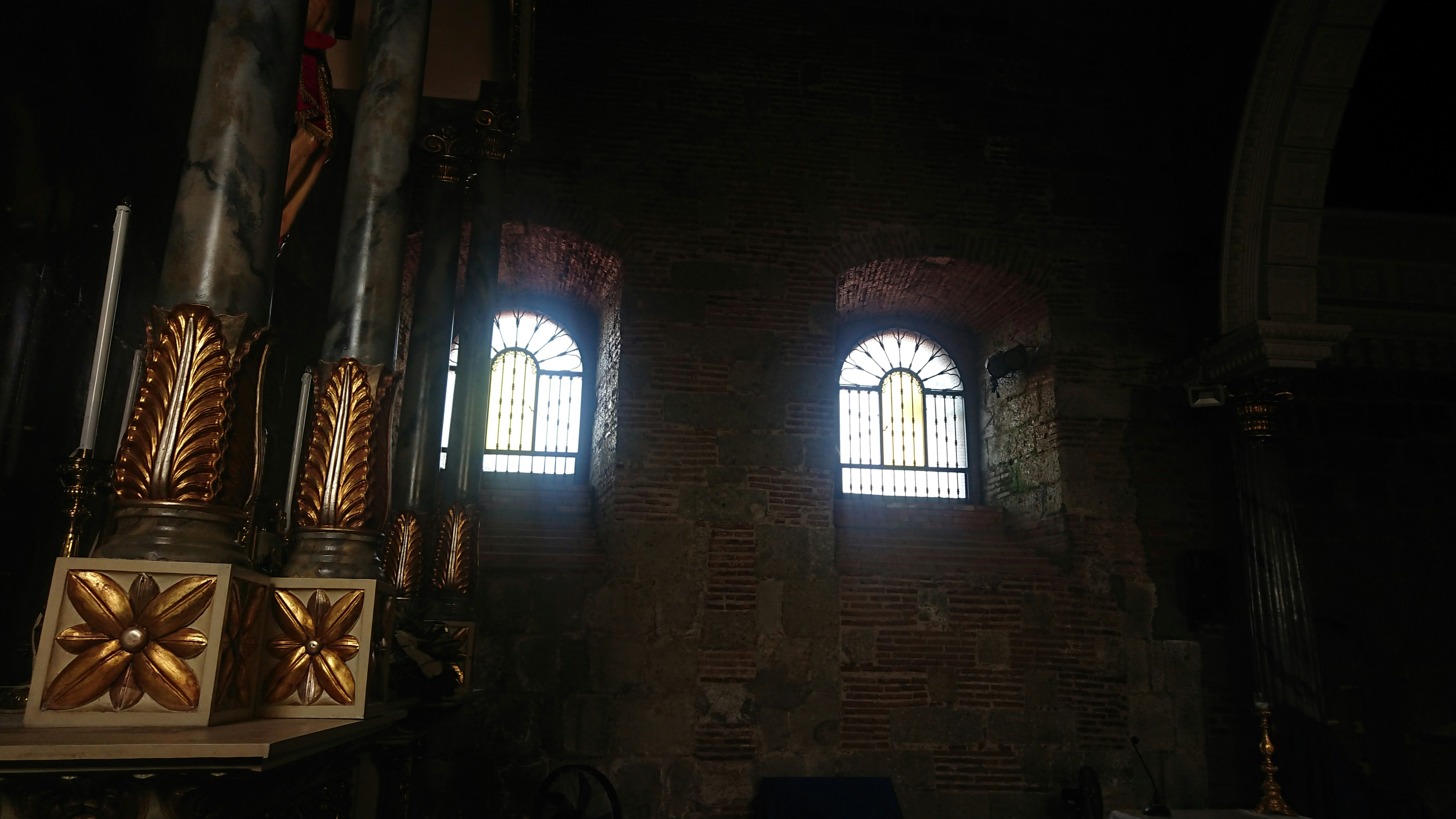 Dim brick nave with two arched windows letting in pale light. Ornamental gold decor rises along a column to the left as the space fades into shadow.