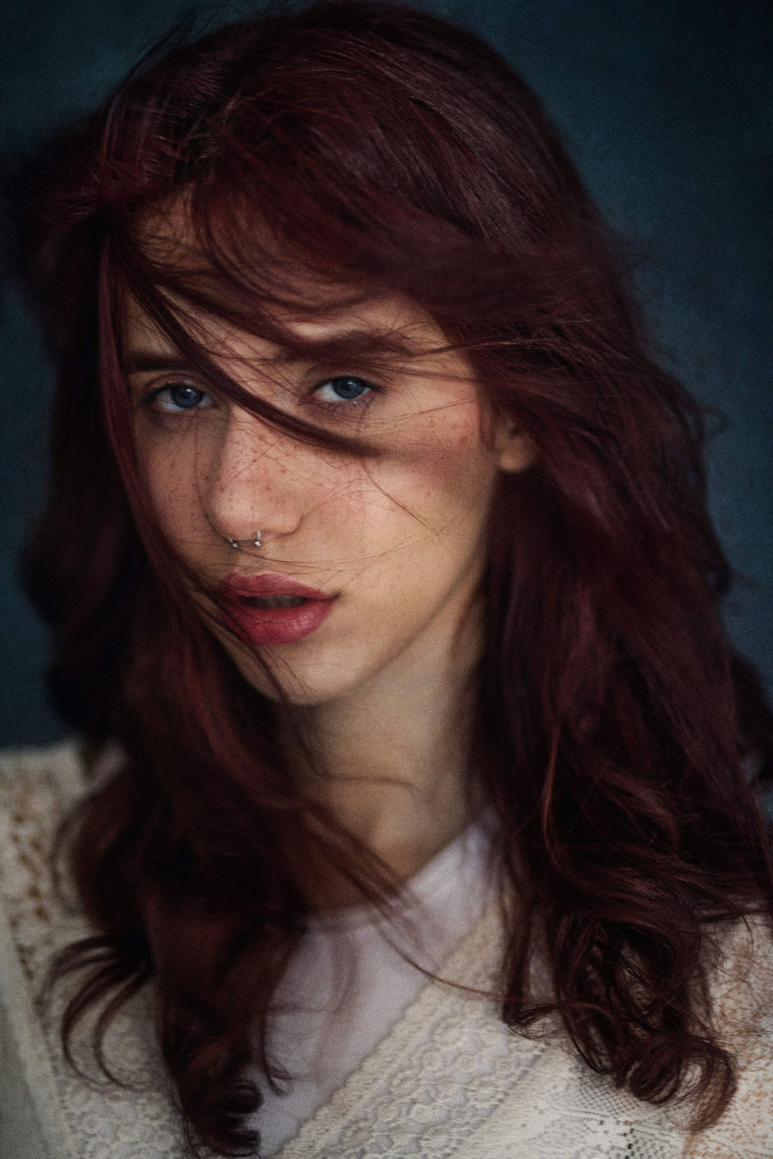 A young woman with flowing red hair and striking blue eyes gazes intently, her hair gently tousled by the breeze. The soft background enhances her features.