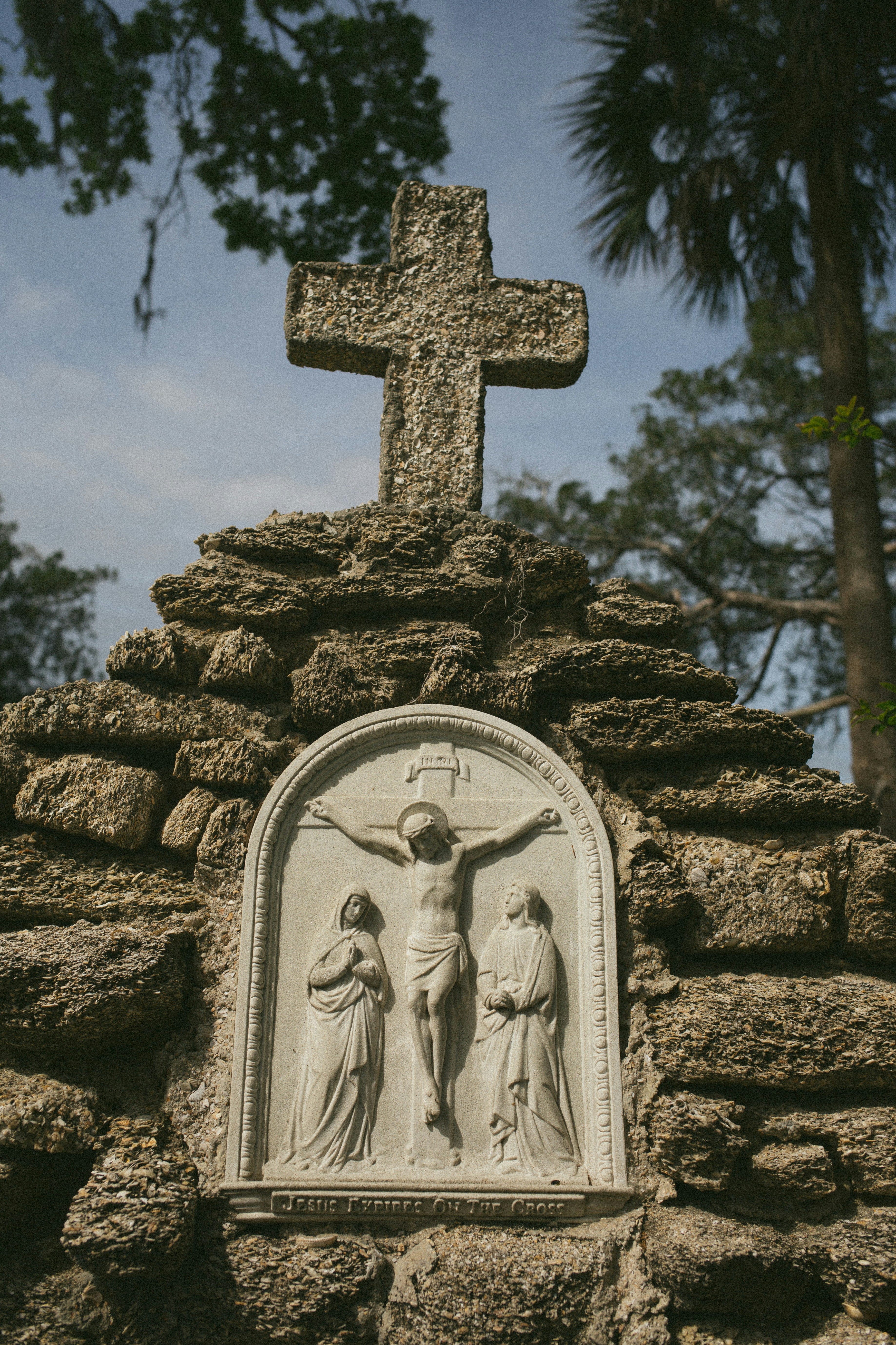 Religious relief sculpture depicting the crucifixion of Jesus, flanked by two figures, set against a rustic stone backdrop.