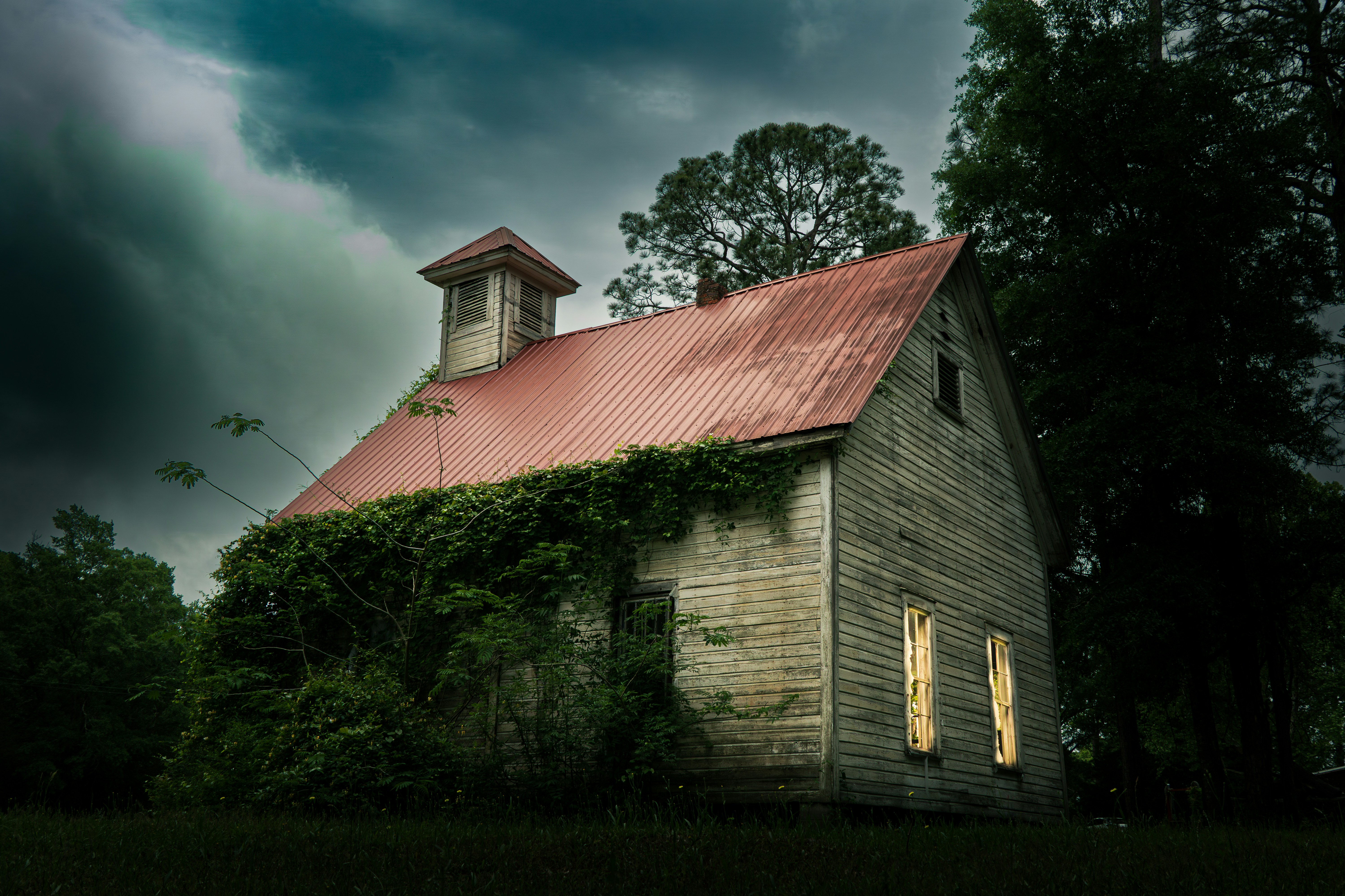 An abandoned house stands alone under a dark, stormy sky. Overgrown vines crawl up its weathered walls, while eerie light glows faintly from the broken windows. Surrounded by dense trees and heavy clouds, the scene feels haunting, mysterious, and full of forgotten stories.