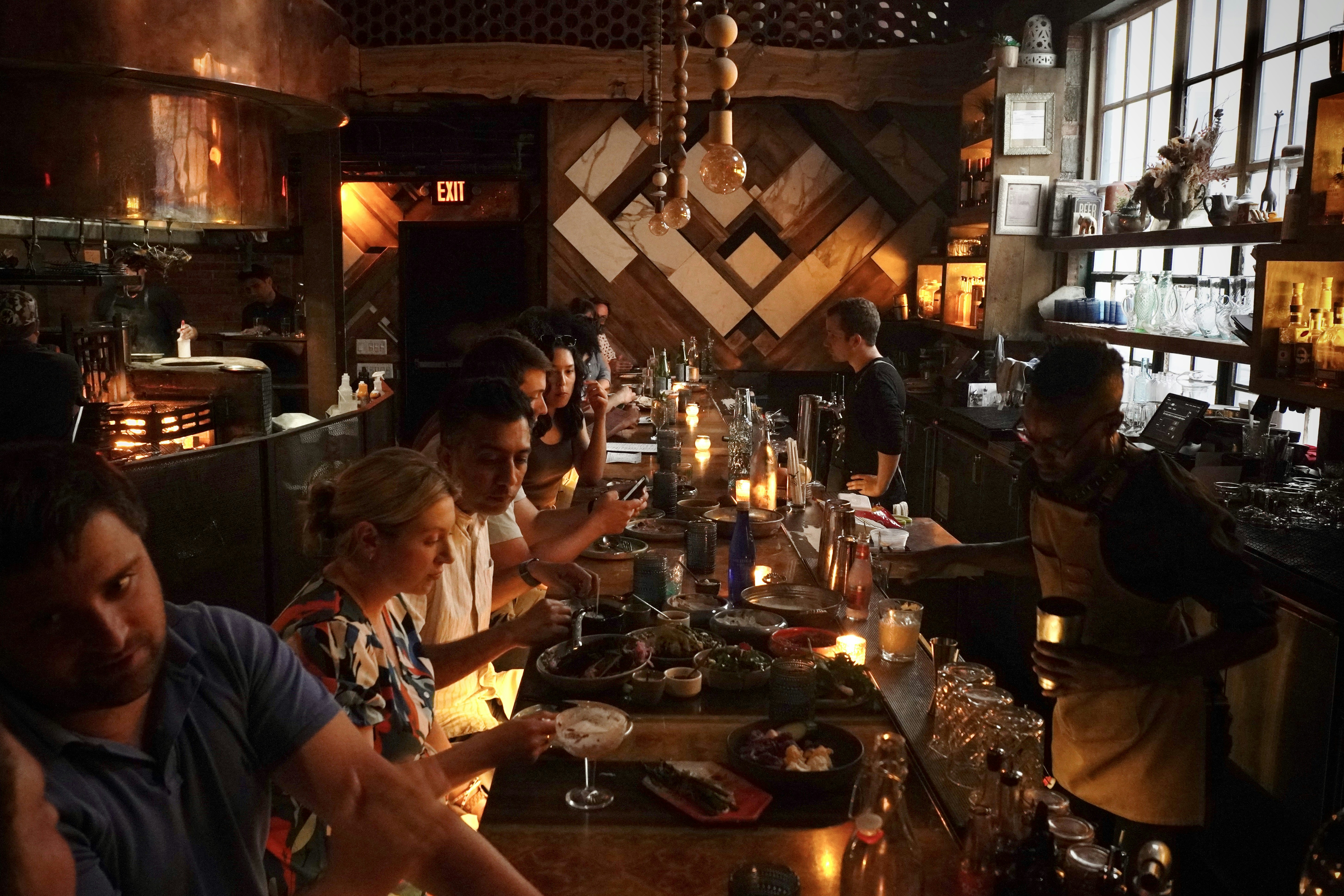 A bustling bar scene with patrons enjoying cocktails and small plates, illuminated by warm candlelight. The bartender skillfully prepares drinks amidst a cozy atmosphere.