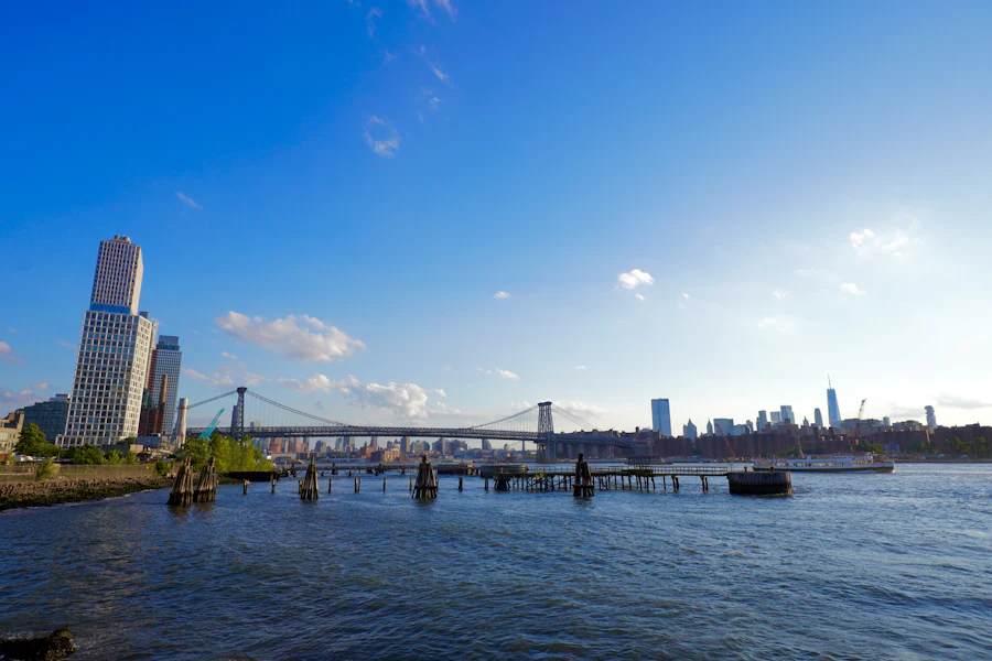 Wide river view with a suspension bridge spanning between a tall riverside tower and a distant city skyline under a clear blue sky.