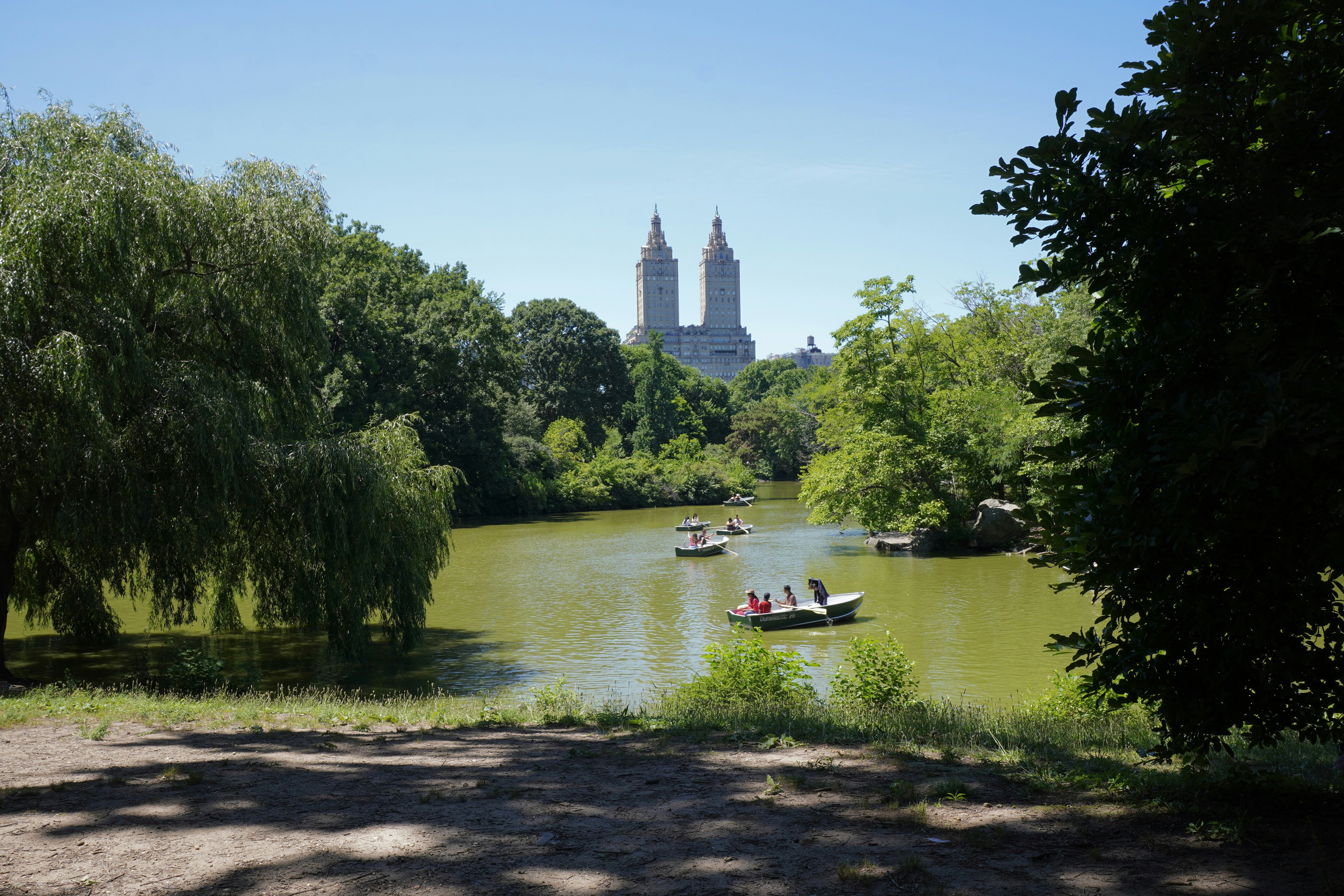 Several rowboats glide peacefully across a calm green lake framed by dense summer foliage. Sunlight filters through the trees, casting soft shadows on the dirt path in the foreground. In the distance, the twin towers of the San Remo rise above the treetops, anchoring the skyline with their iconic silhouette. The scene captures a relaxed summer afternoon in the heart of nature and the city.