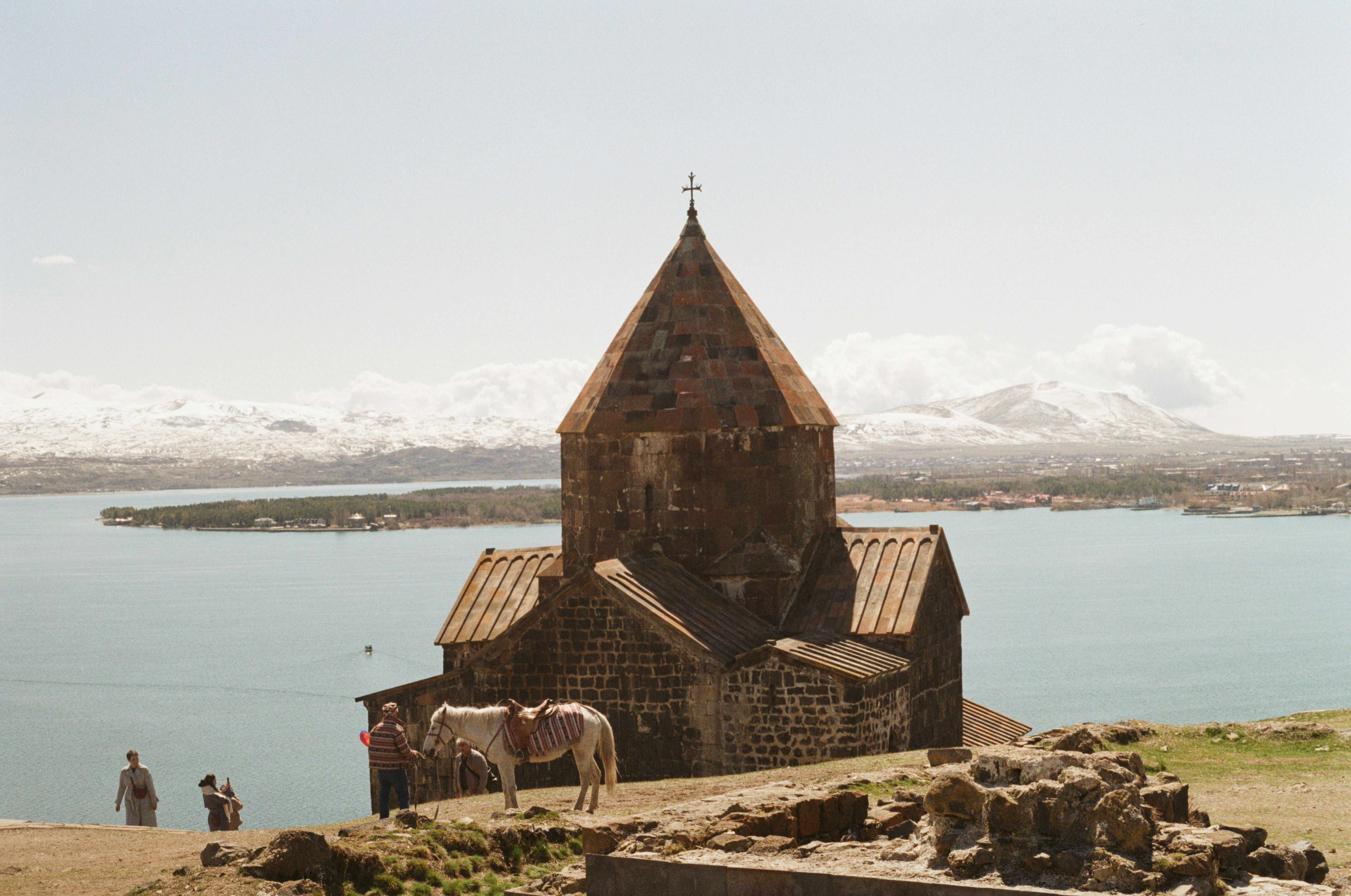 Stone chapel on a rocky promontory beside a calm fjord, with a grazing horse and a few figures nearby, and distant snow-capped mountains beyond the shore.