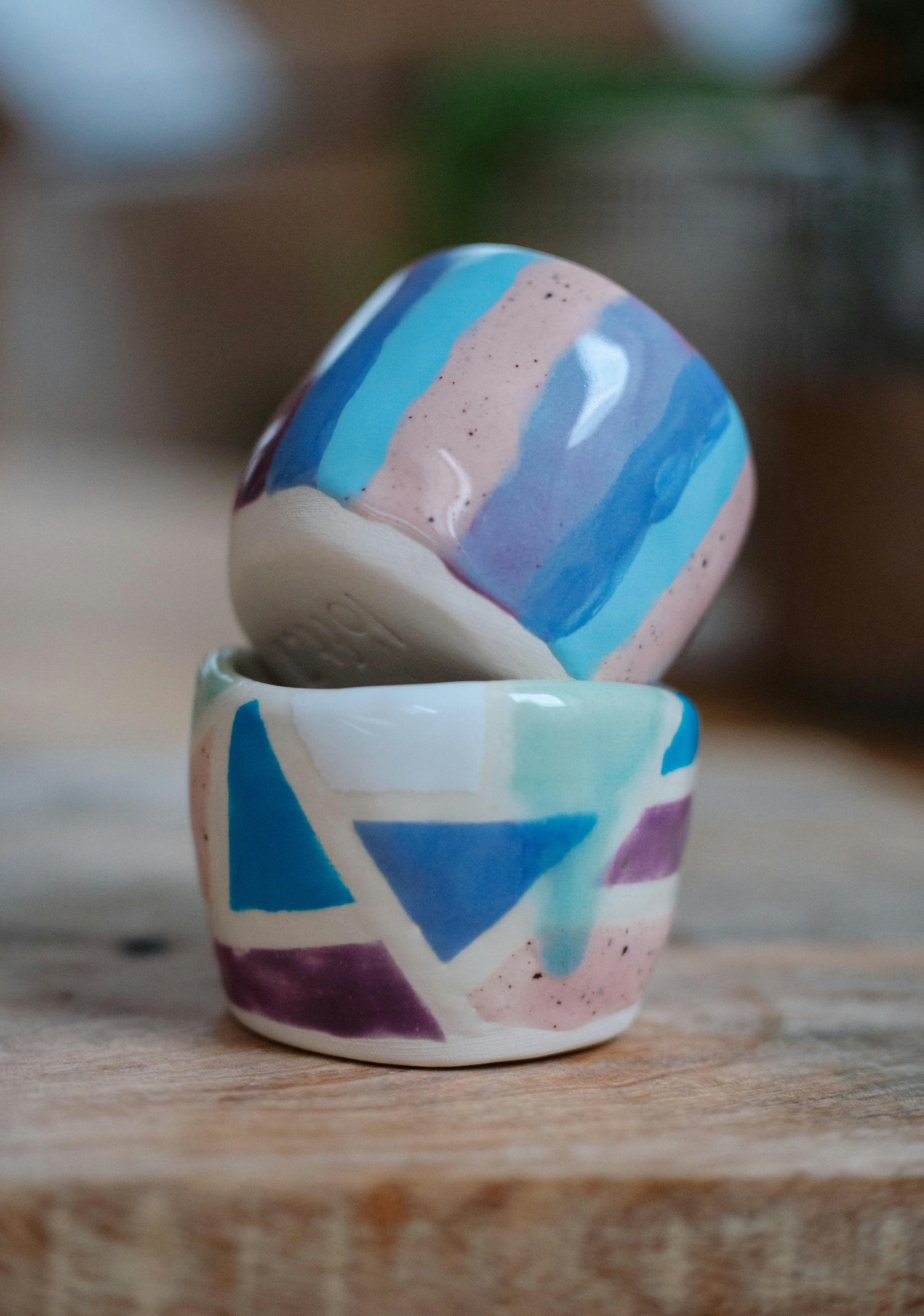 Two uniquely patterned ceramic cups stacked on a wooden surface, showcasing vibrant geometric designs and a blend of pastel colors.