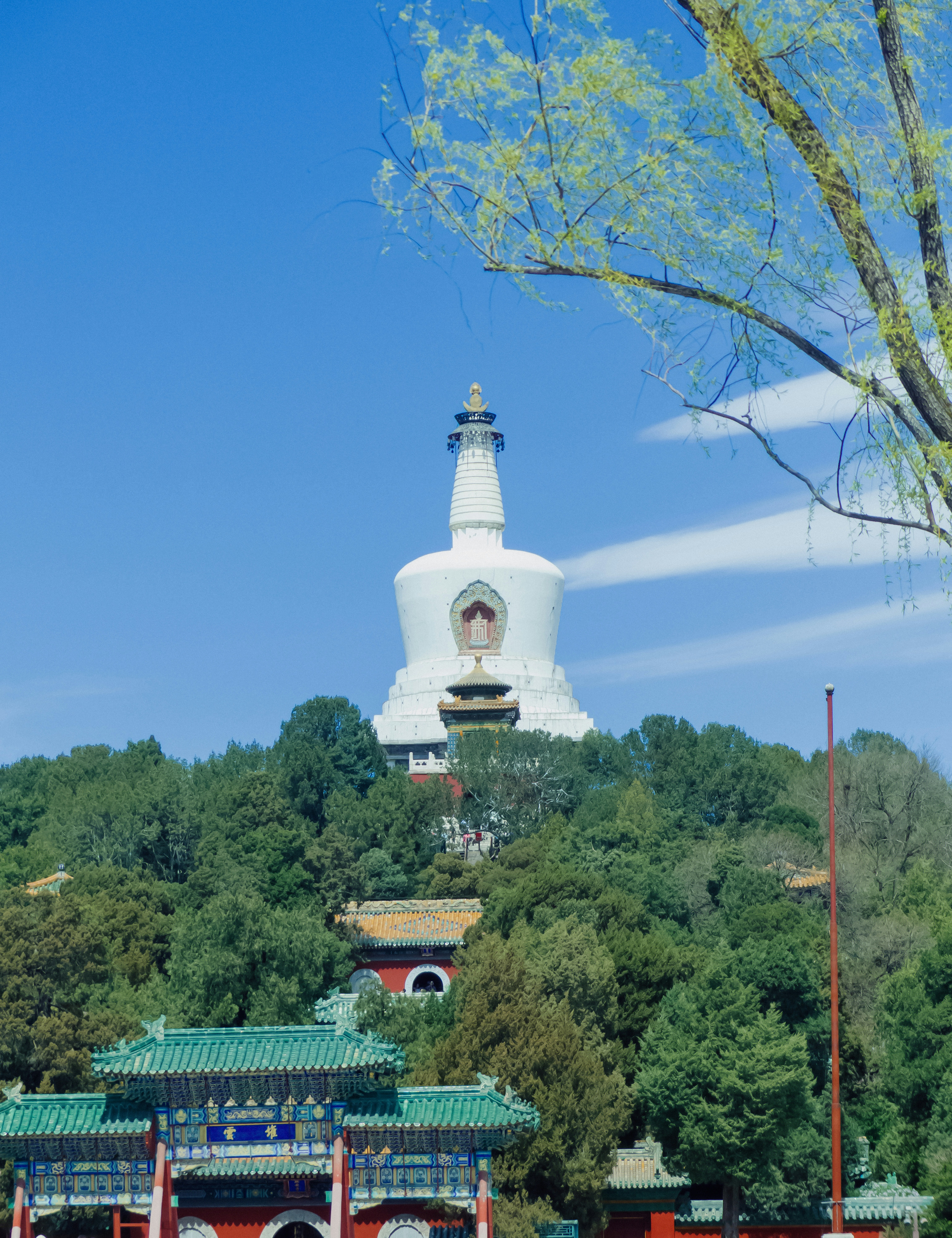 A white stupa perched on a forested hill, framed by a Chinese temple complex with teal roofs and a bright blue sky.