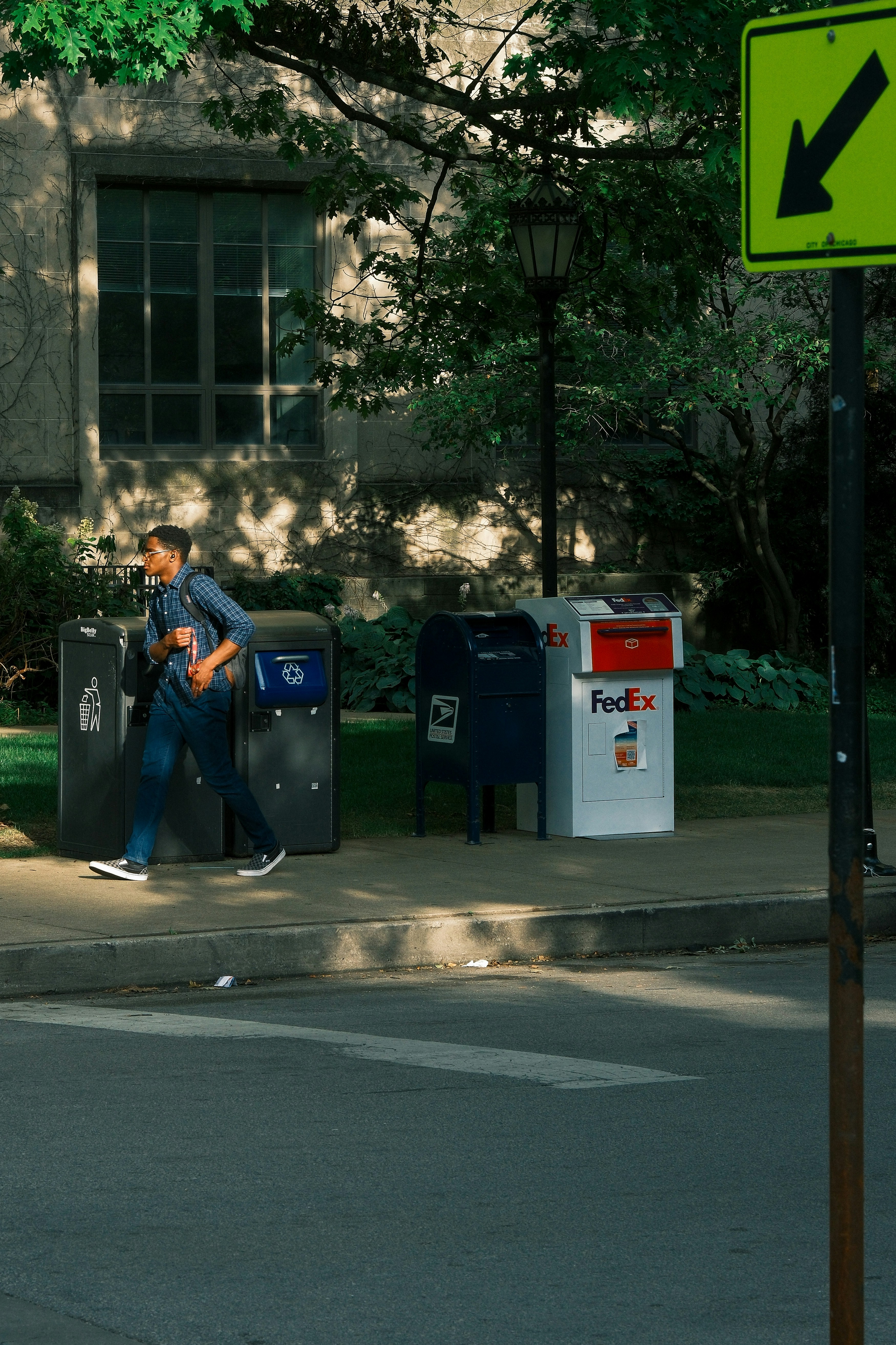 A person walks past a row of mailboxes, including a FedEx box, under dappled sunlight in an urban setting.