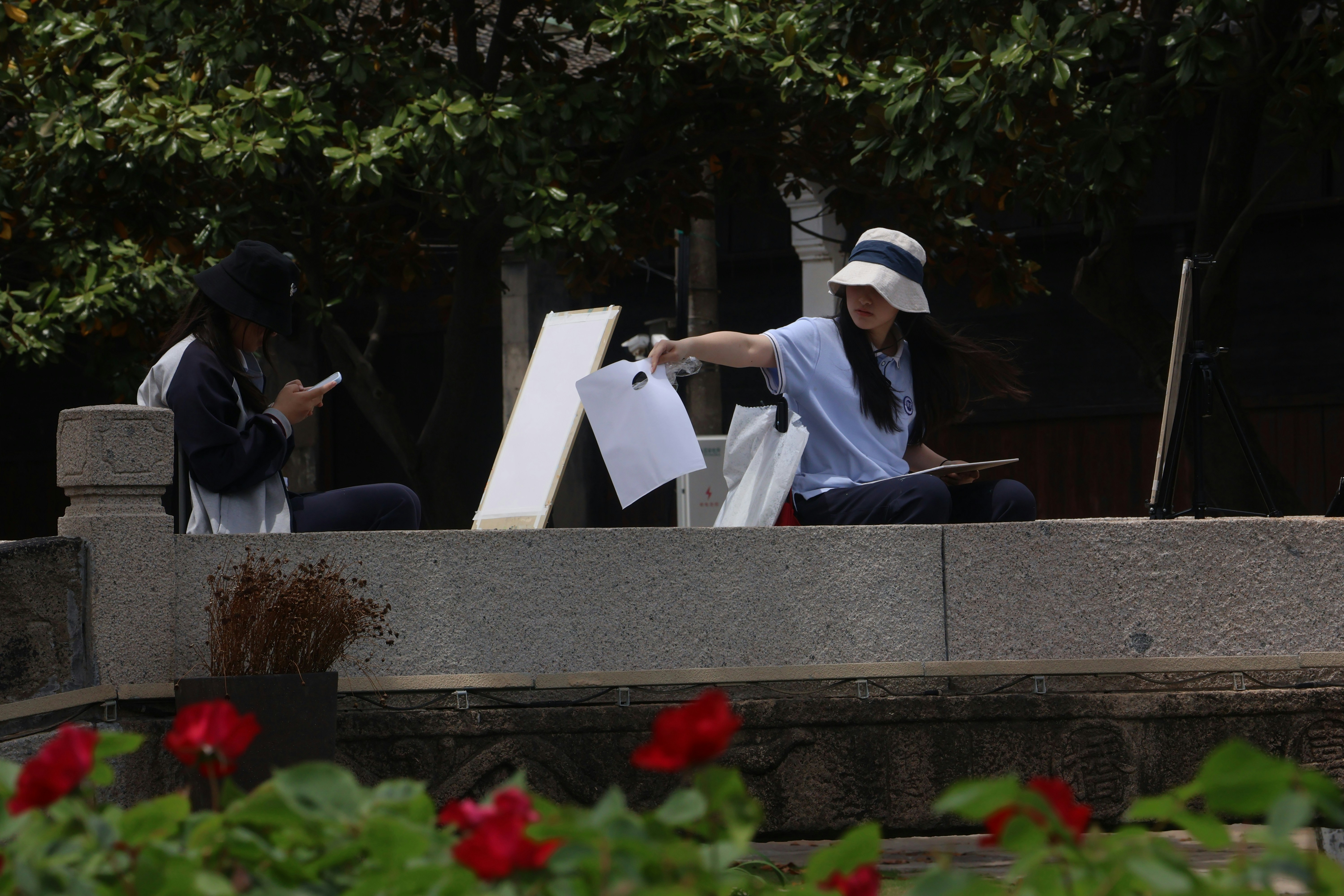 Two individuals engaged in creative work by a serene water feature, surrounded by vibrant flowers. The scene captures a moment of artistic endeavor in a lush environment.