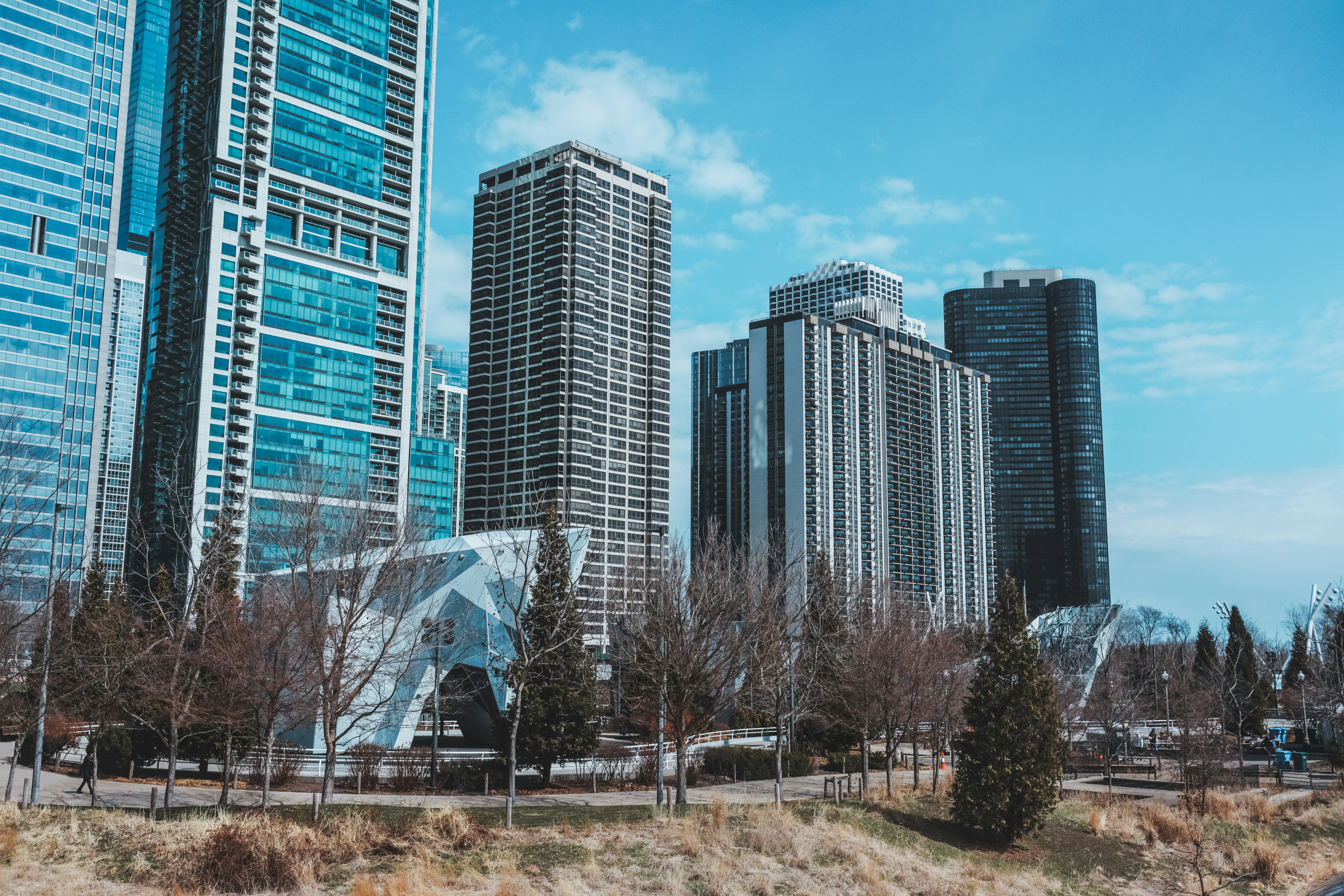 Winter cityscape with tall glass skyscrapers rising behind a small park featuring a white geometric sculpture and leafless trees.