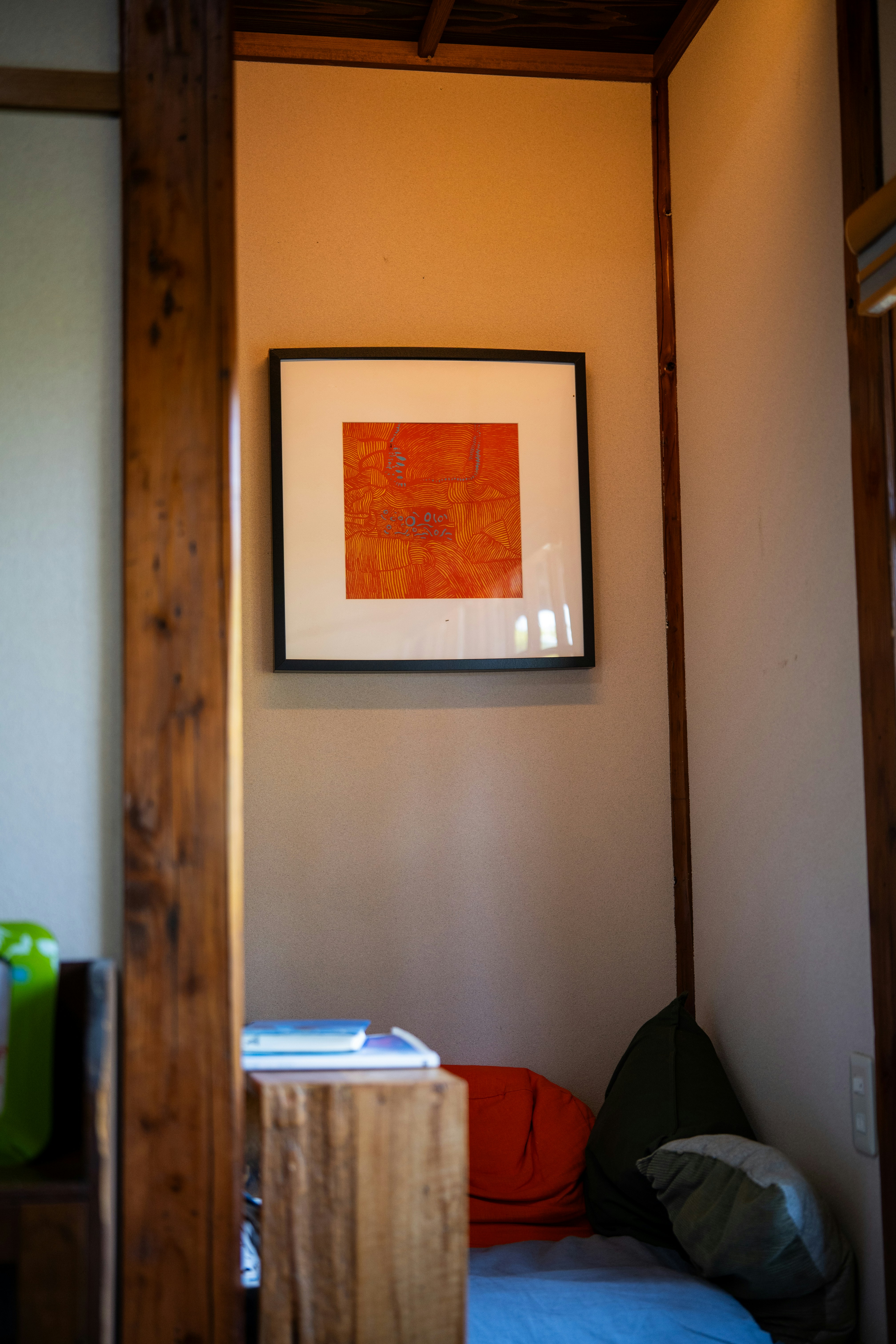 Framed orange abstract painting hangs on a beige wall within a timber-framed alcove, with a small furniture cluster and cushions below.