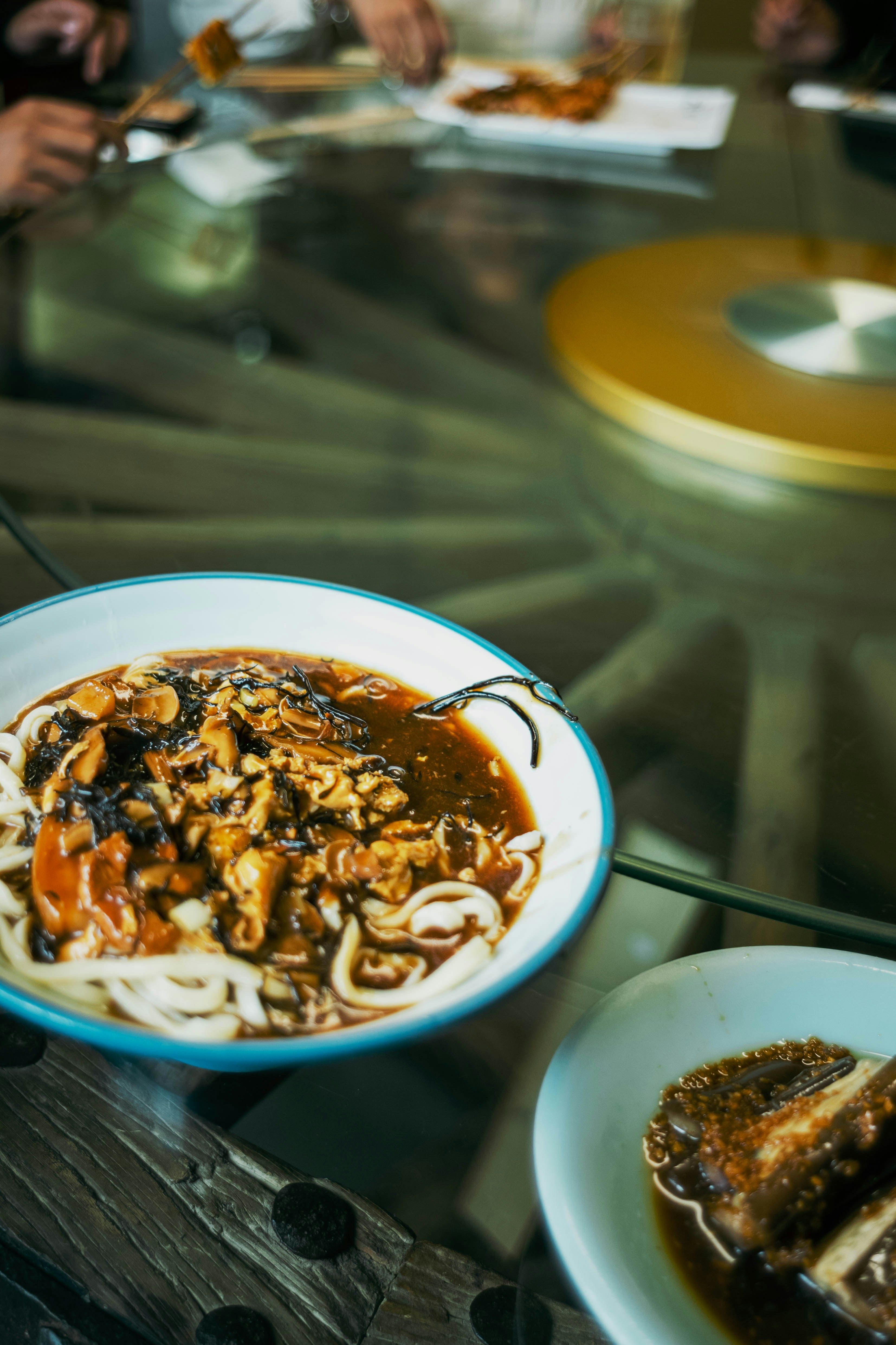 Umami in Focus: Noodle Bowl on a Glass Table Amid Blur of Tabletop Conversation