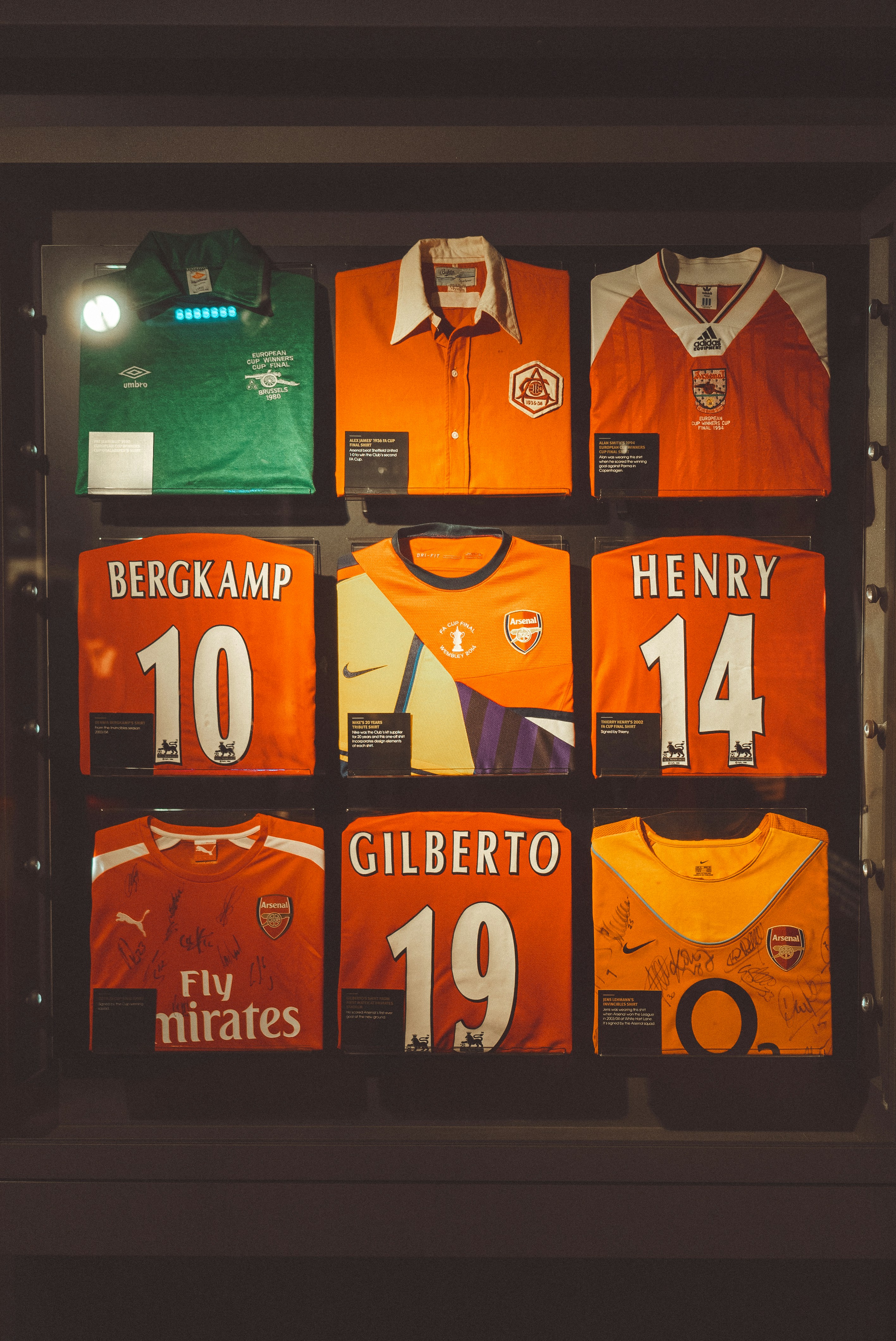 Nine Arsenal football jerseys are arranged in a 3x3 grid inside a glass display case. Each jersey features a visible name and number.
