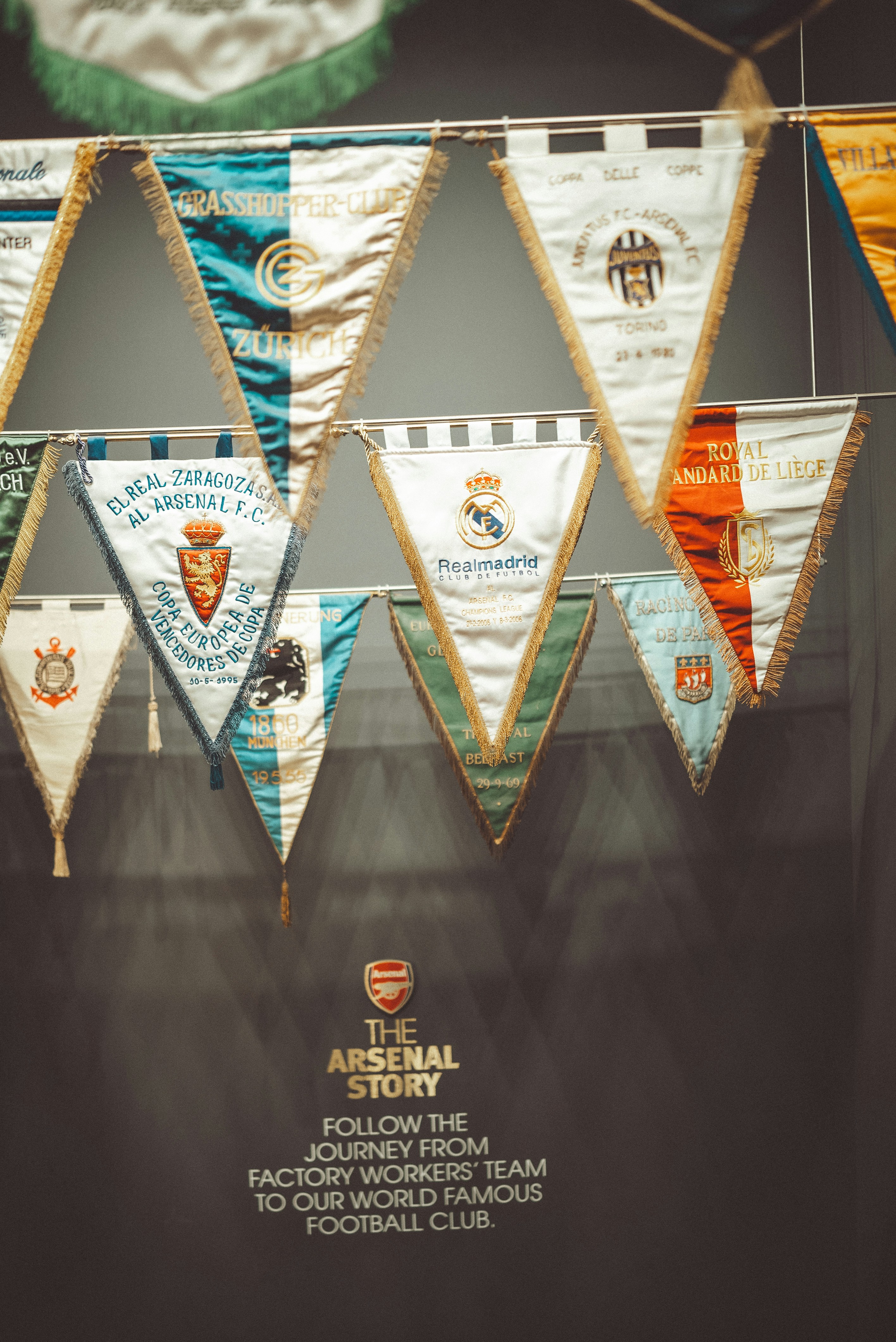 Colorful pennants representing various football clubs hang against a dark background, showcasing the rich history of the sport. Below, a plaque narrates the story of a factory workers' team evolving into a renowned football club.
