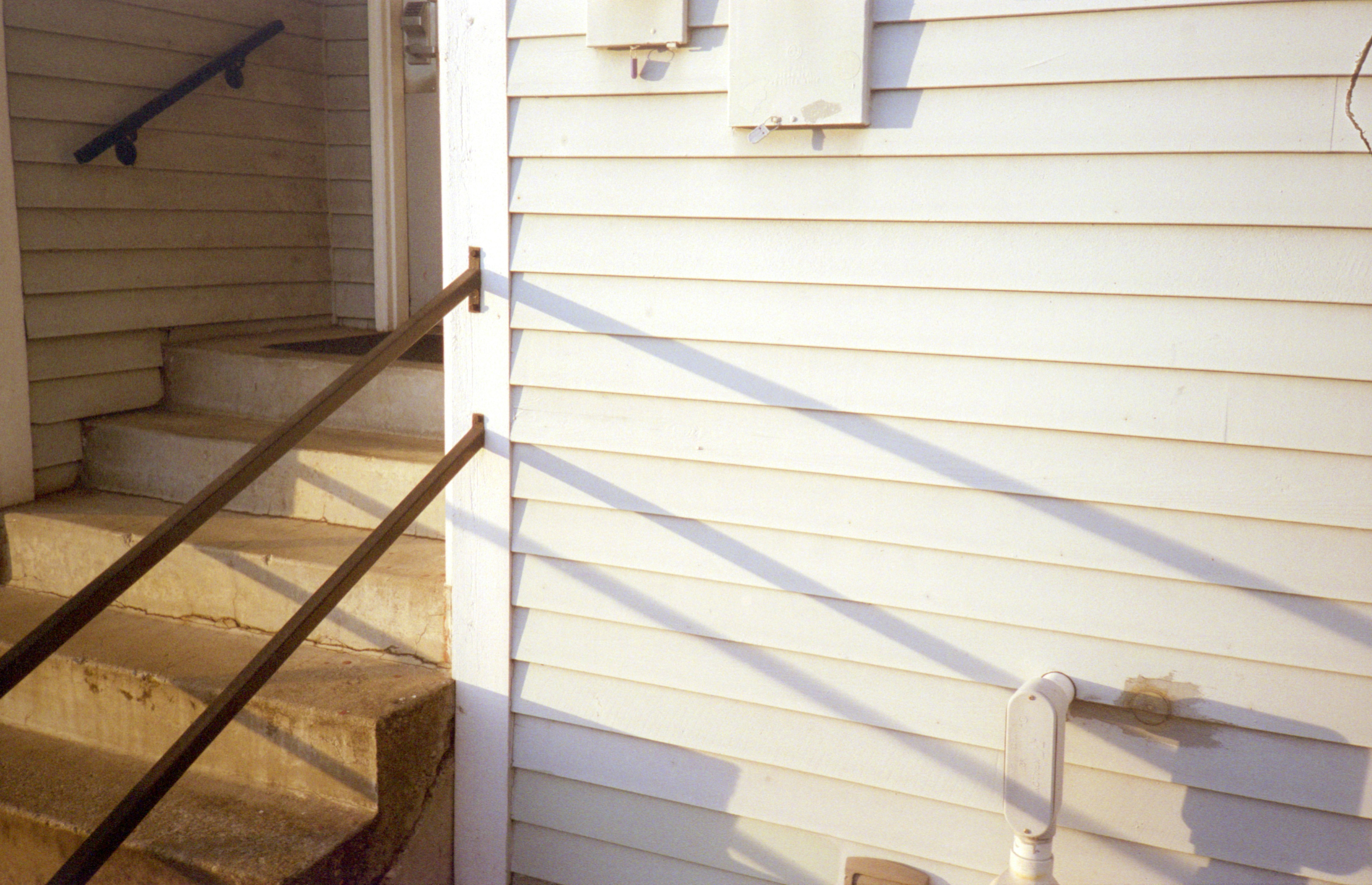 Exterior wall with pale siding, a metal handrail along concrete steps on the left, and diagonal shadows sweeping across the surface.
